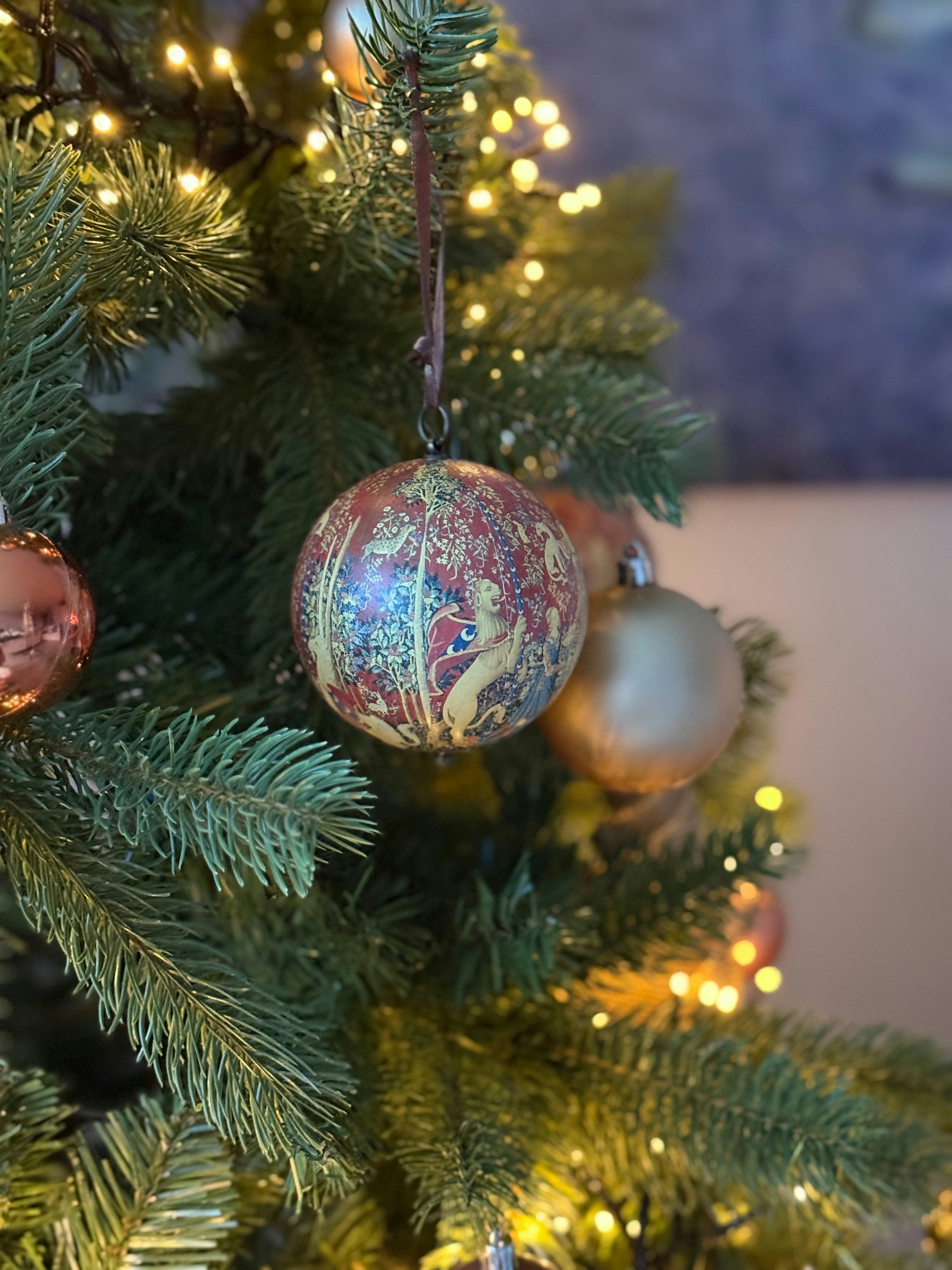 Christmas ornaments on a decorated Christmas tree. The main ornament displays a design inspired by the medieval Flemish tapestries known as ‘The Lady and the Unicorn’ (French: La Dame à la licorne), which symbolically represents ‘taste’ and ‘smell’ in the mille-fleurs style. This artwork is housed at the Musée de Cluny in Paris, France.