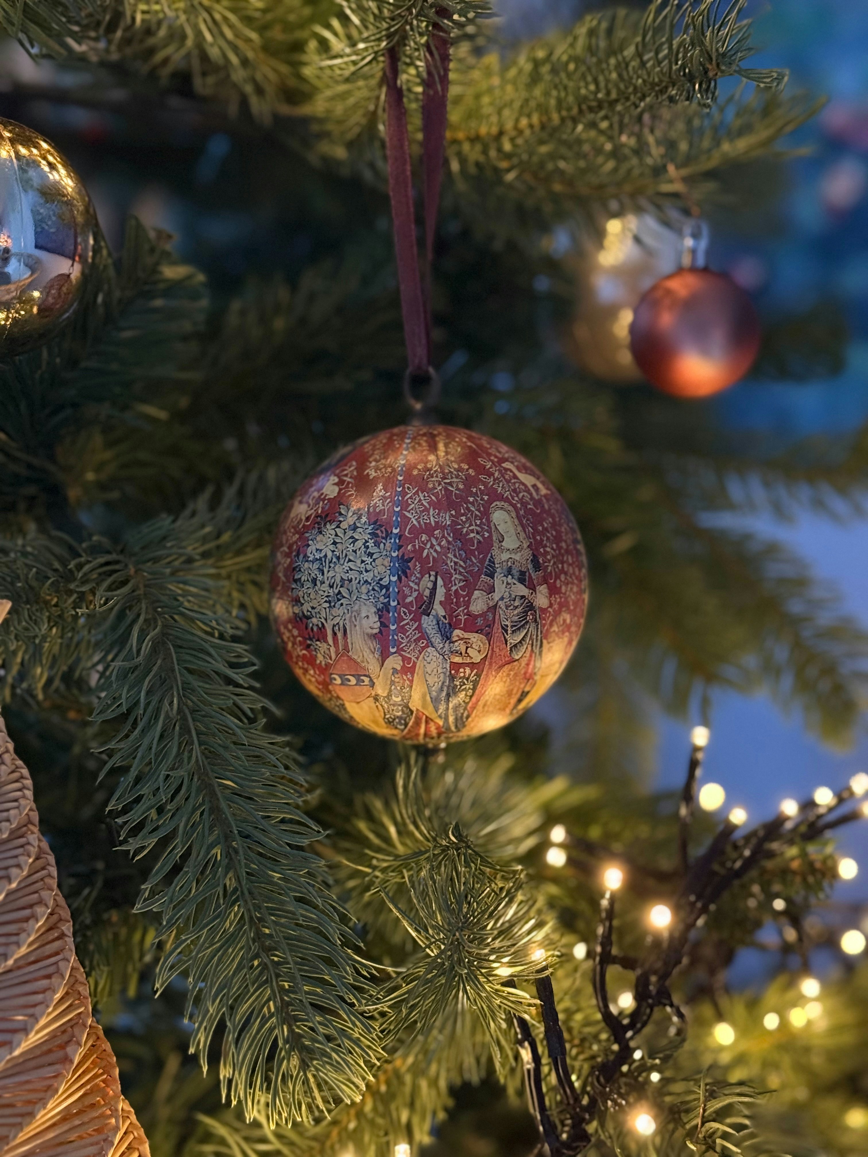 Christmas ornaments on a decorated Christmas tree. The main ornament displays a design inspired by the medieval Flemish tapestries known as ‘The Lady and the Unicorn’ (French: La Dame à la licorne), which symbolically represents ‘taste’ and ‘smell’ in the mille-fleurs style. This artwork is housed at the Musée de Cluny in Paris, France.