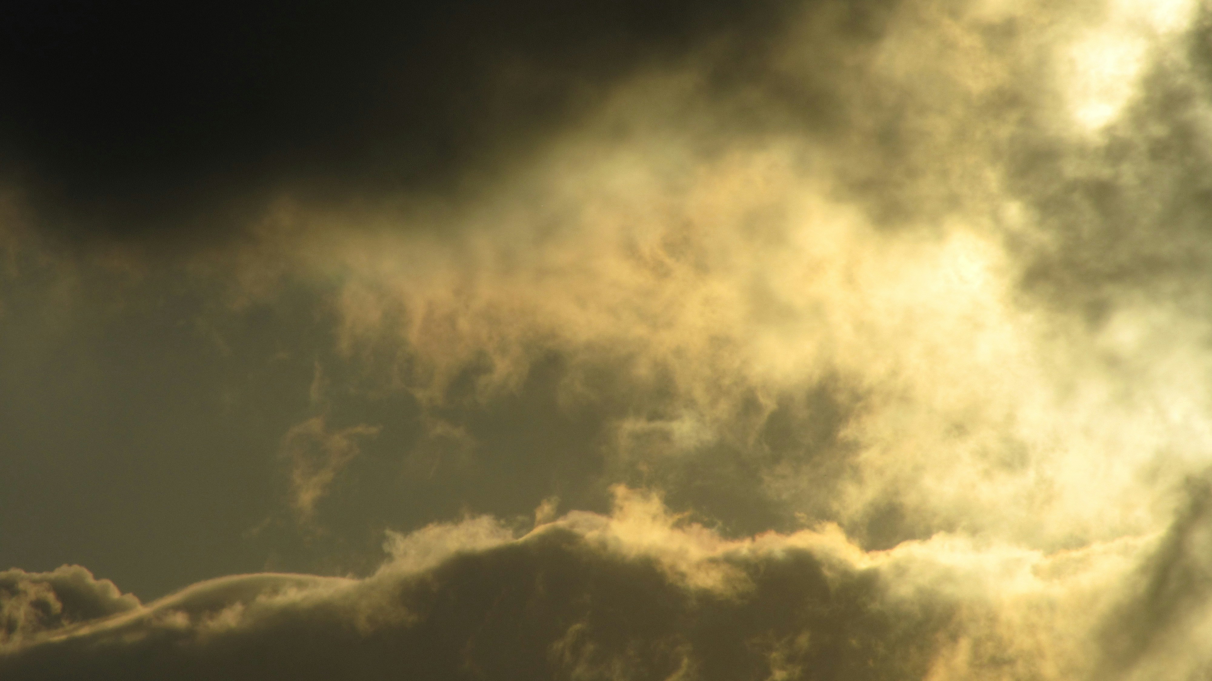 A moody sky where heavy storm clouds meet soft golden sunlight. The light breaks through the haze, highlighting the swirling textures of the clouds and creating a dramatic, cinematic atmosphere.