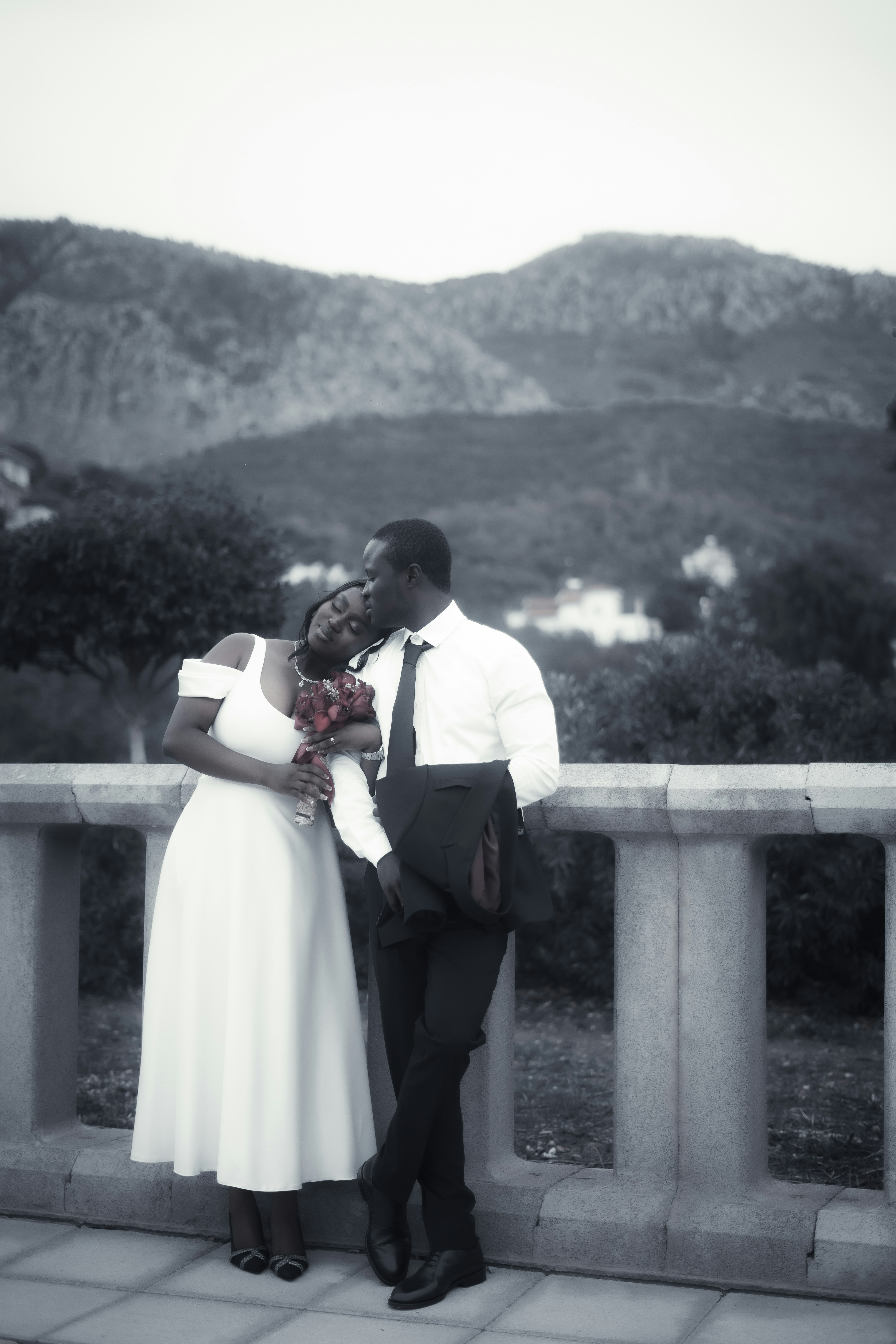 A tender moment between a couple captured against a dramatic mountain backdrop. The woman rests peacefully on her partner’s shoulder, holding a bouquet of roses, while he leans in with a gentle kiss. The soft, muted tones and serene landscape add an intimate, heartfelt mood to the scene, highlighting the closeness and calm of their connection.