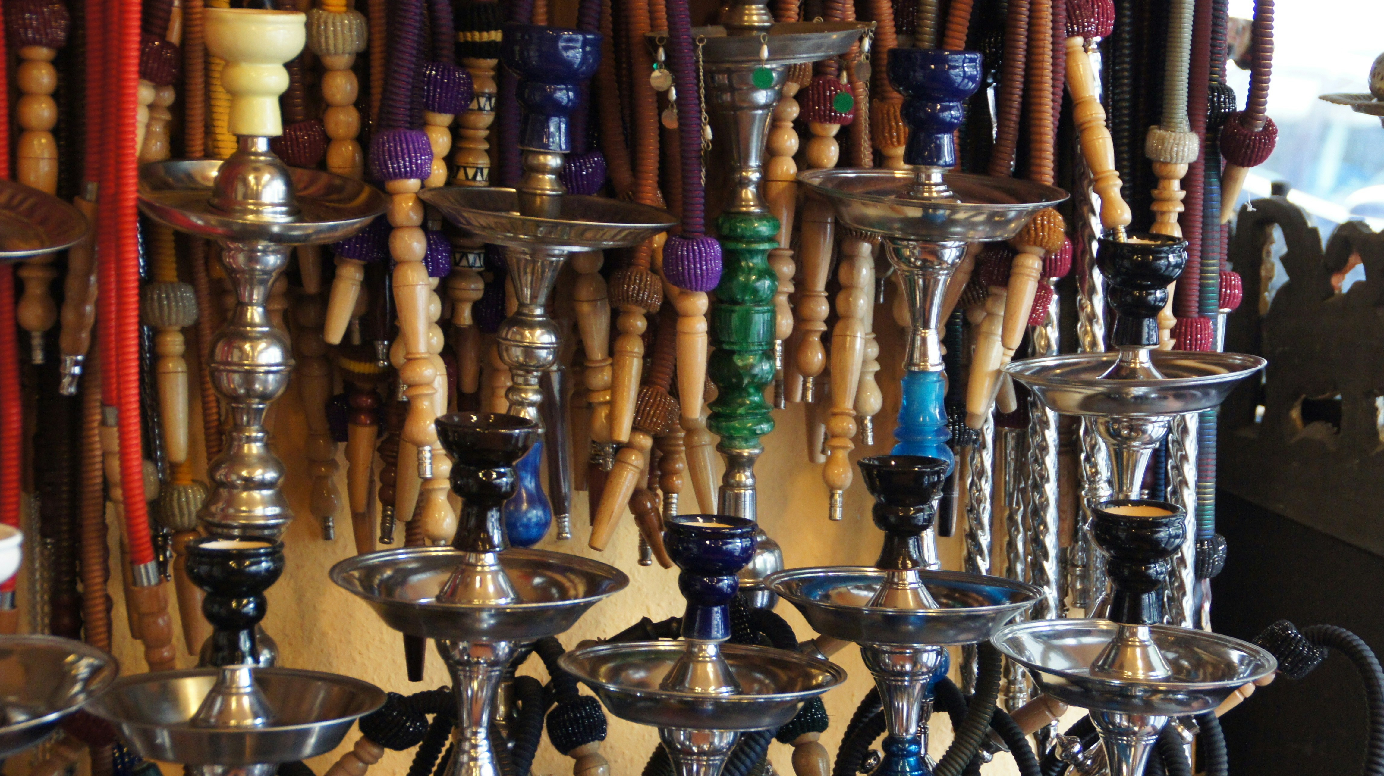 A colorful display of hookahs and mouthpieces fills the frame. The mix of wood, metal, and vibrant colors creates a lively market atmosphere. The objects are tightly arranged, forming a textured and dynamic composition. Reflections on the metal surfaces add depth and shine. The image captures the charm of a traditional hookah shop.