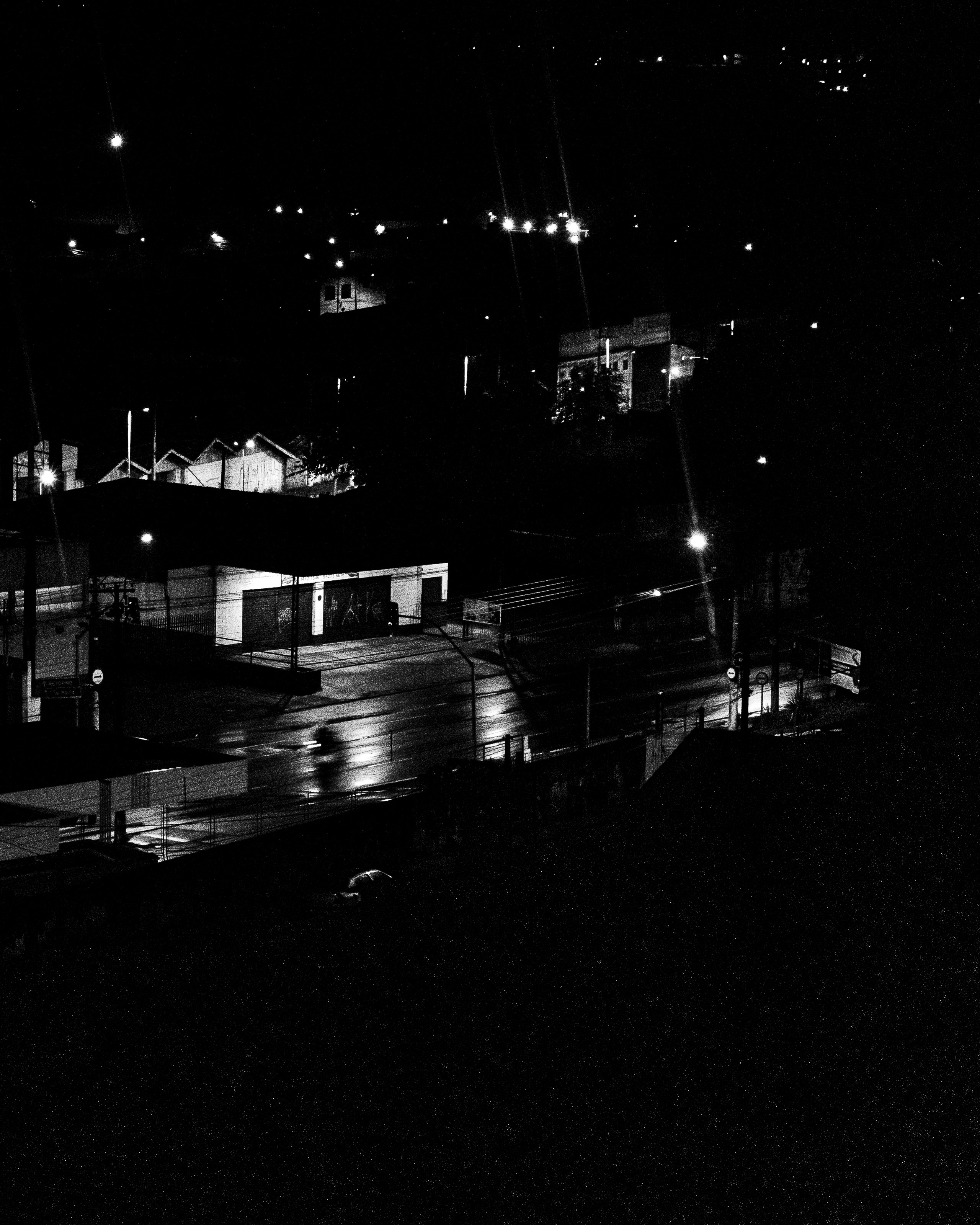 A blurry figure on a motorcycle drives down a wet street at night, illuminated by streetlights and the lights of buildings.