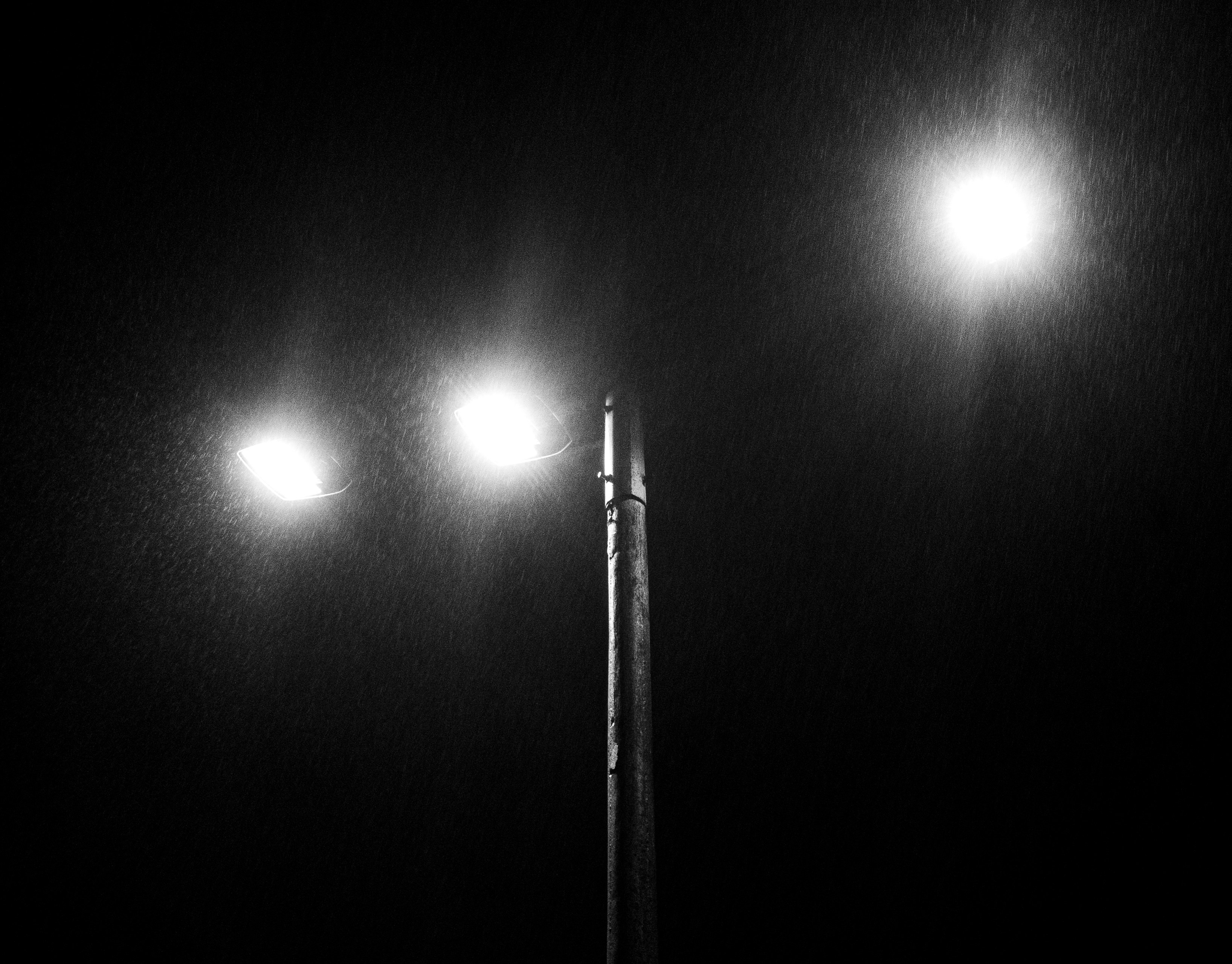 A street lamp with three lights shines brightly in the dark, with rain falling around it.