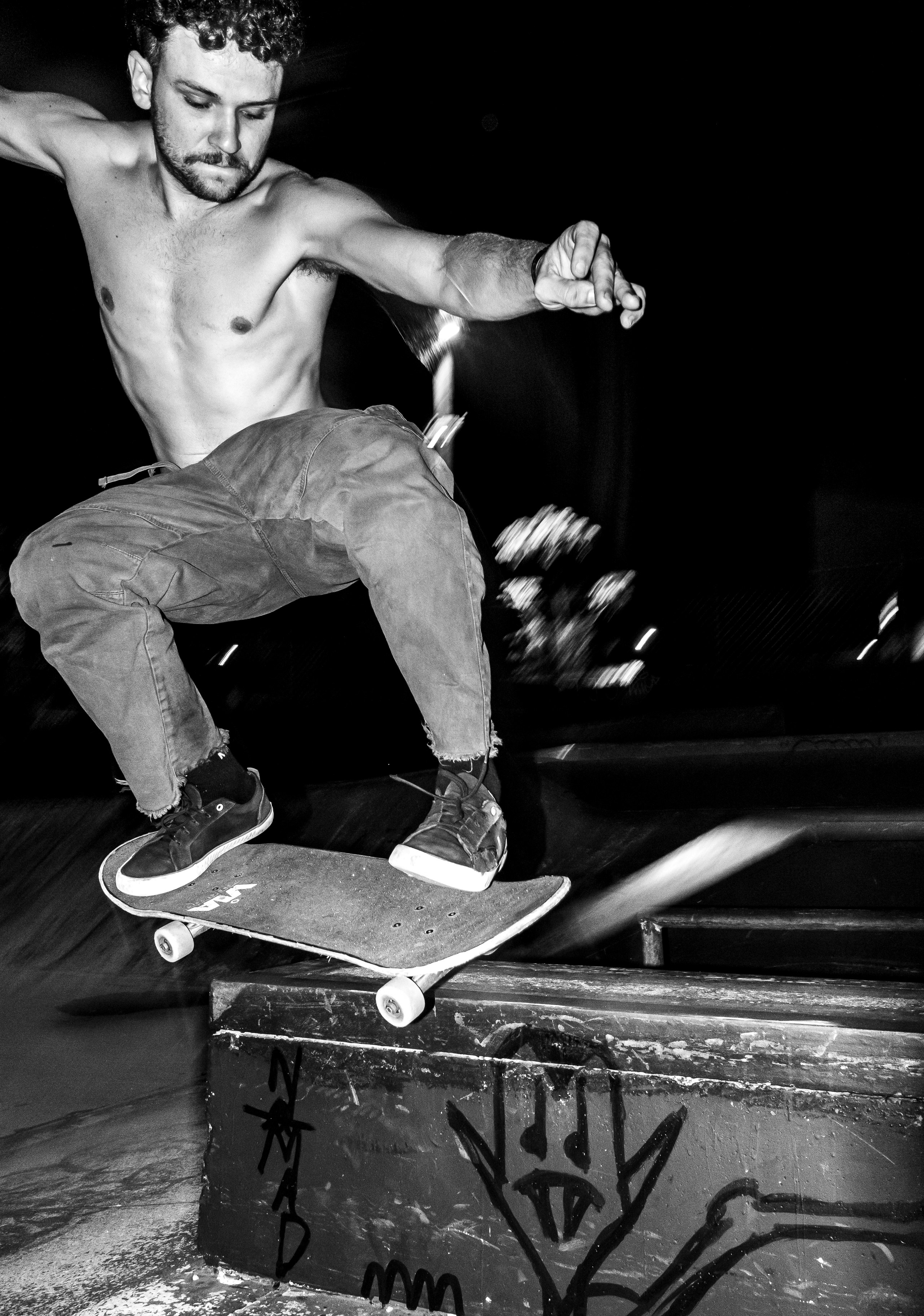 A shirtless man with curly hair and a mustache performs a skateboard trick at night, jumping over a ledge in a skatepark.