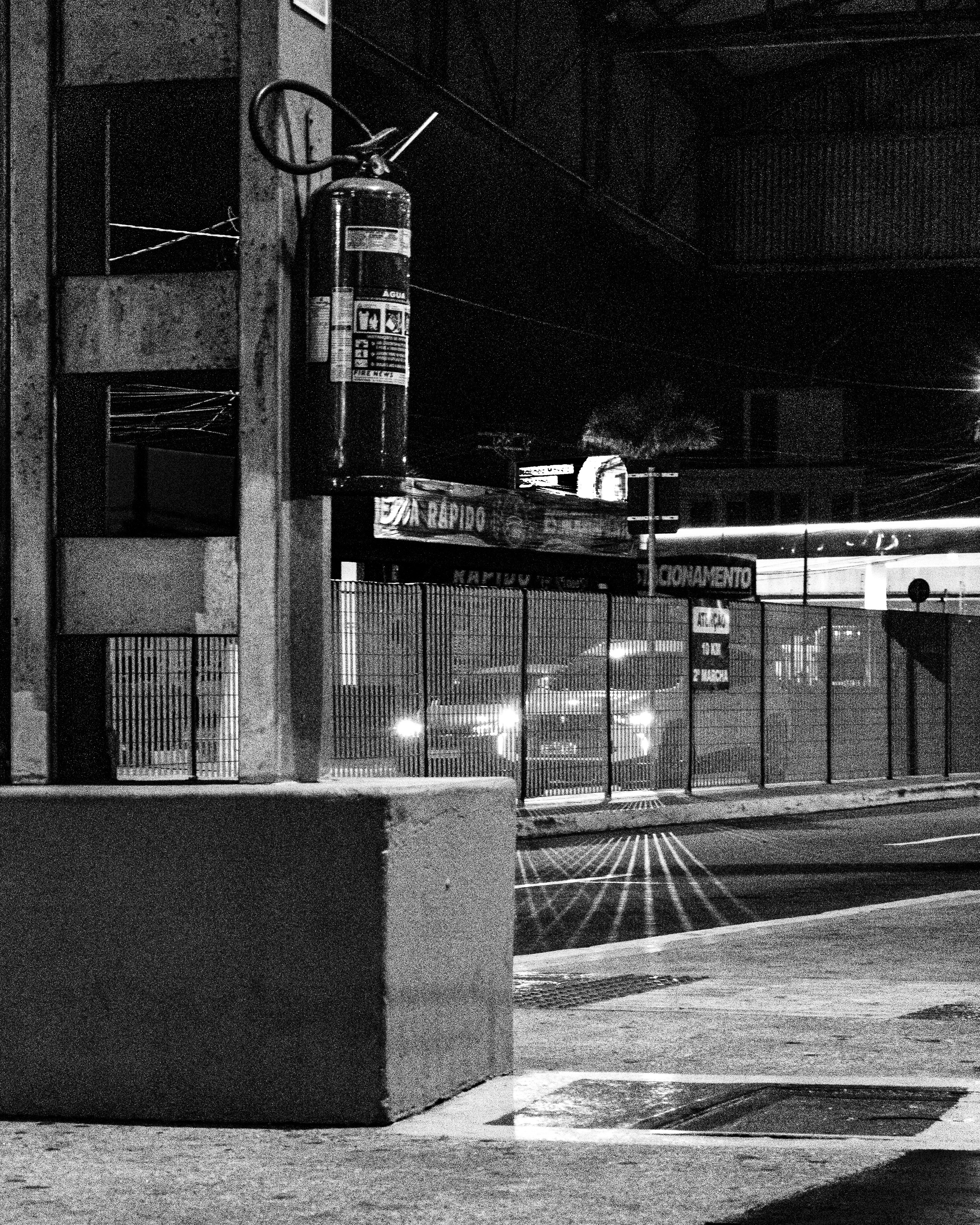 A fire extinguisher hangs from a pole in front of a building at night. The building has a sign that reads "EM RÁPIDO" and another sign that reads "CIONAMENTO". There are cars parked behind a fence in the background.