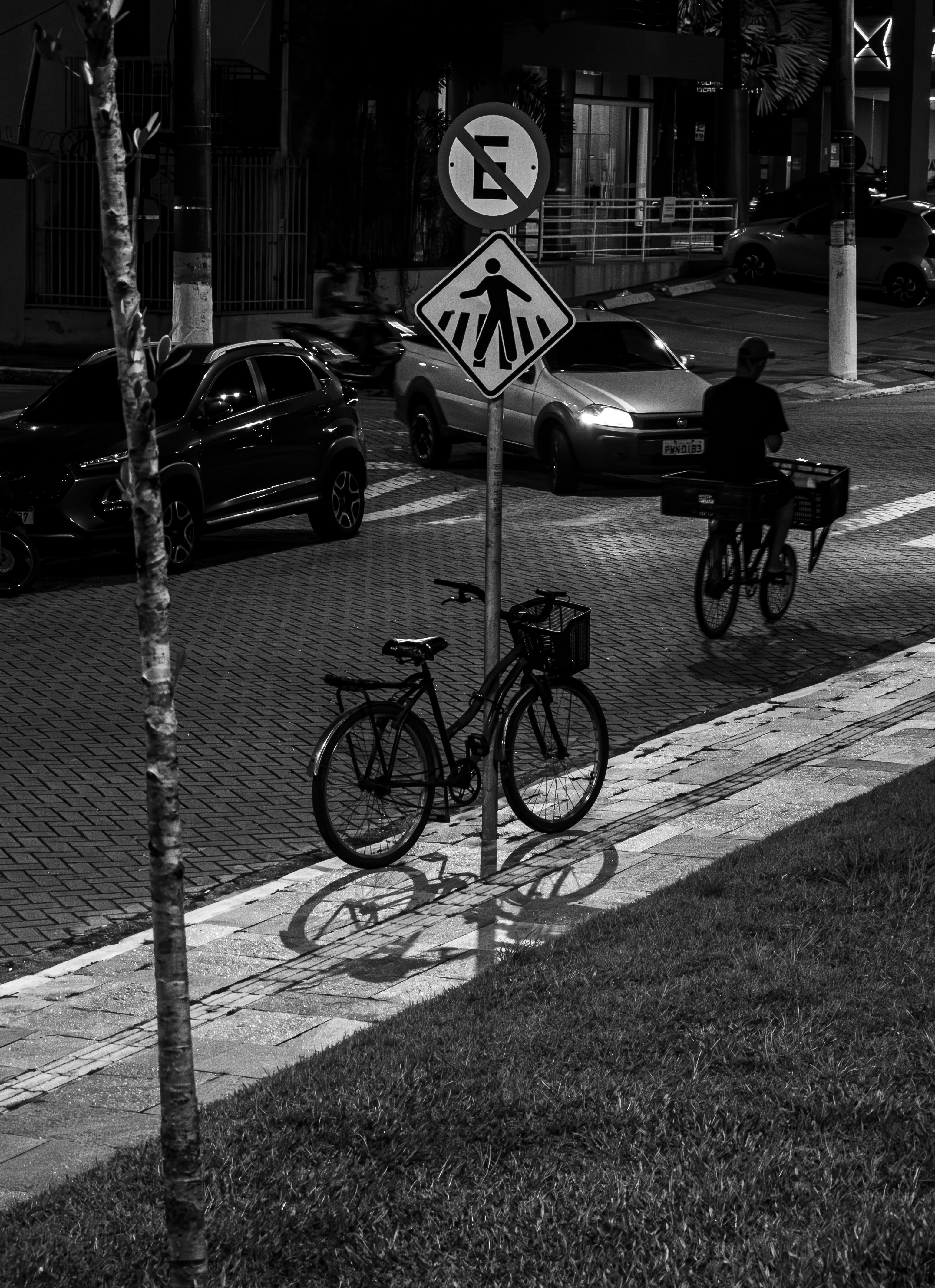 A delivery person on a bicycle with a large basket on the back rides past a pedestrian crossing sign at night.