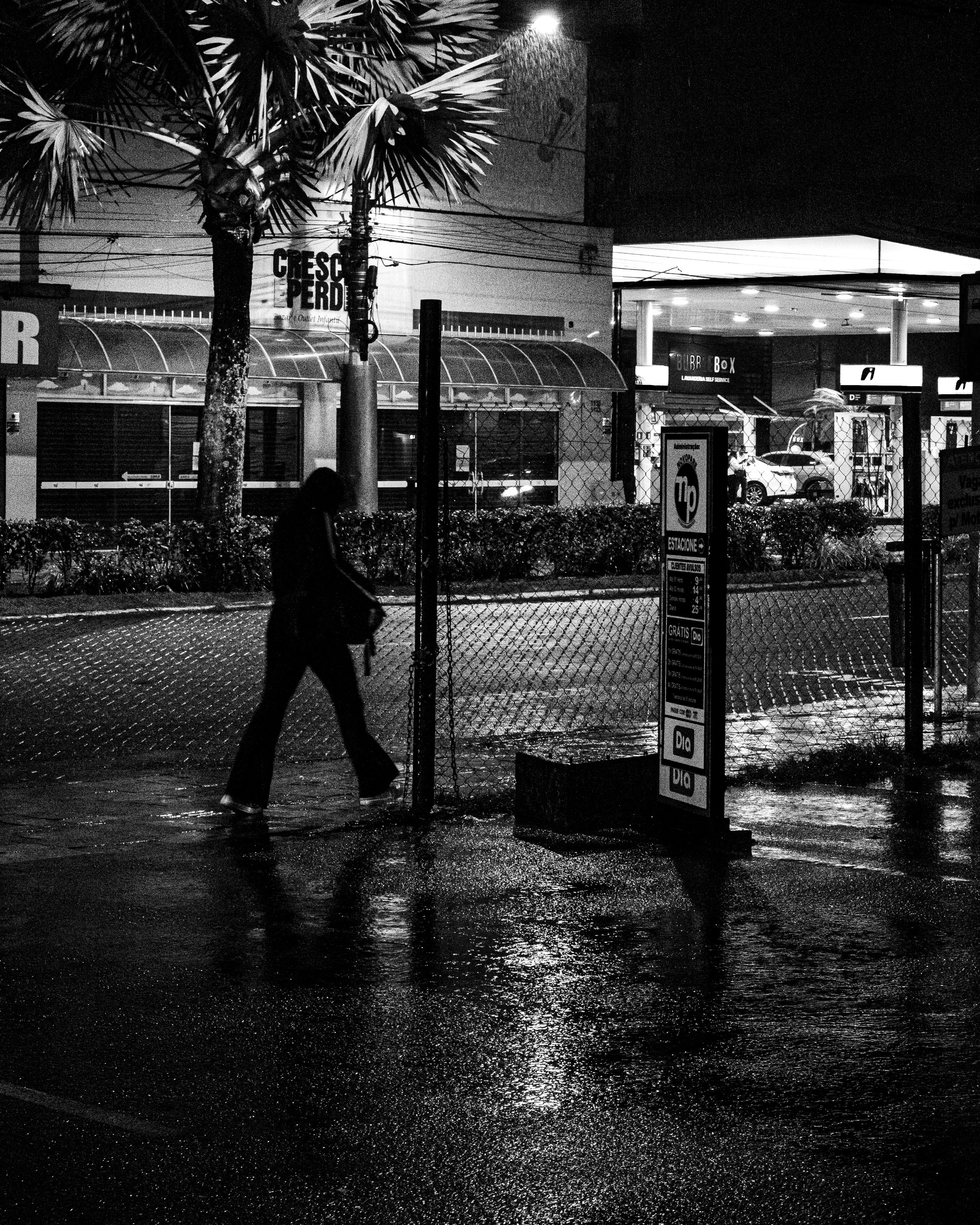 A lone figure walks down a wet street at night, illuminated by the lights of a gas station and a street lamp. The rain creates reflections on the pavement, adding to the moody atmosphere of the scene.