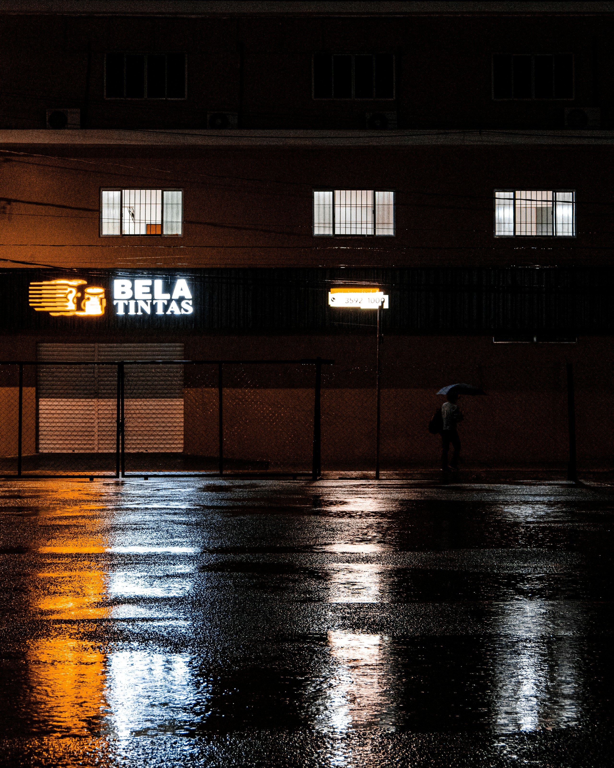 A lone person walks with an umbrella on a wet street at night, illuminated by the neon sign of Bela Tintas and reflections from streetlights.