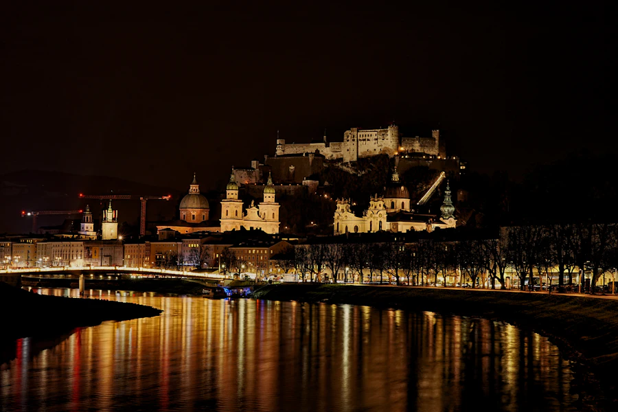 Nighttime cityscape along a river with illuminated historic buildings and a fortress perched atop a hill; reflections shimmer on calm water, conveying a tranquil, nocturnal atmosphere.