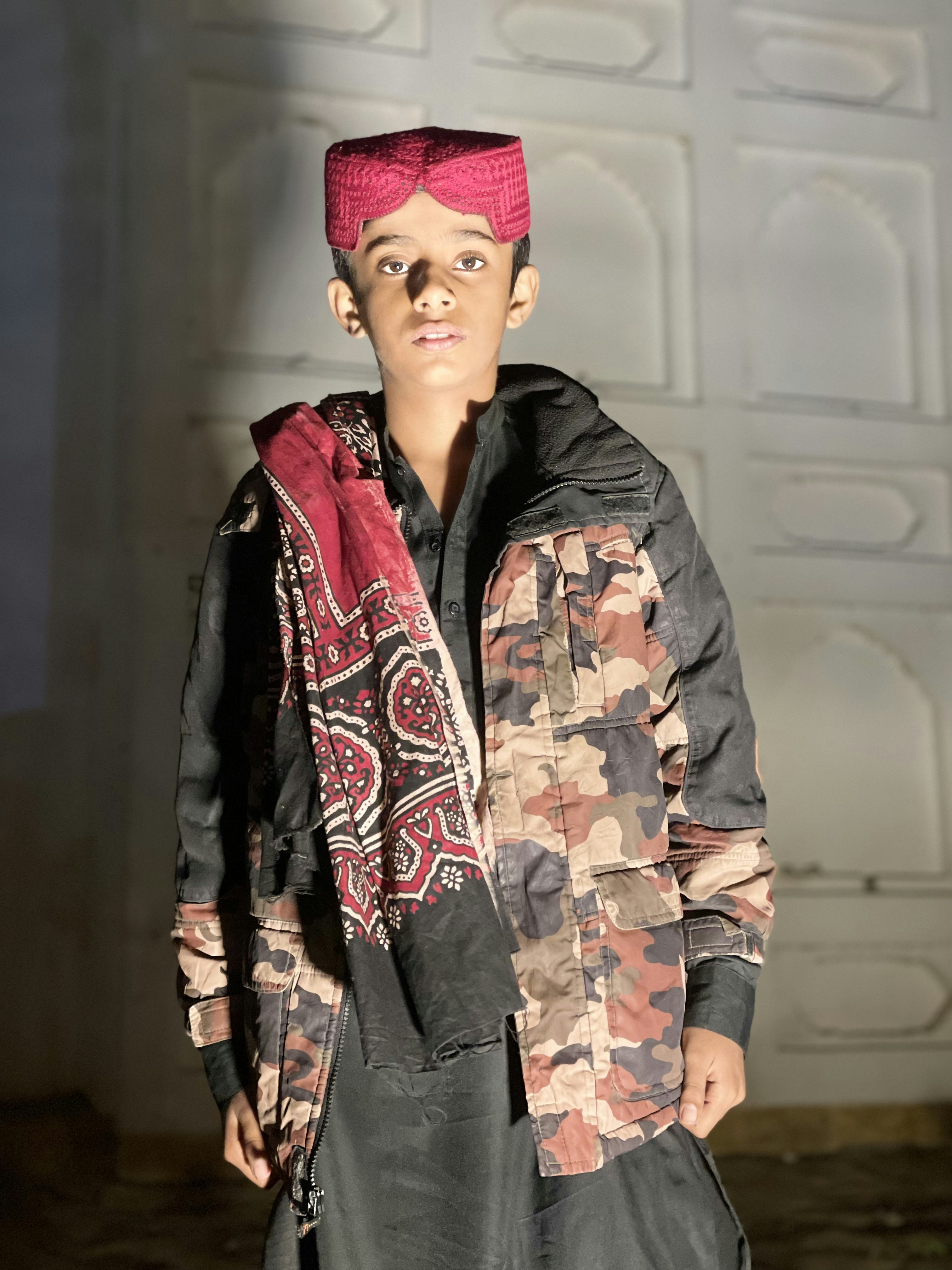 Young man in a bold red turban and layered camouflage-patterned jacket stands before a decorative wall, illuminated from the front. A fashion-forward portrait emphasizing texture and cultural attire.