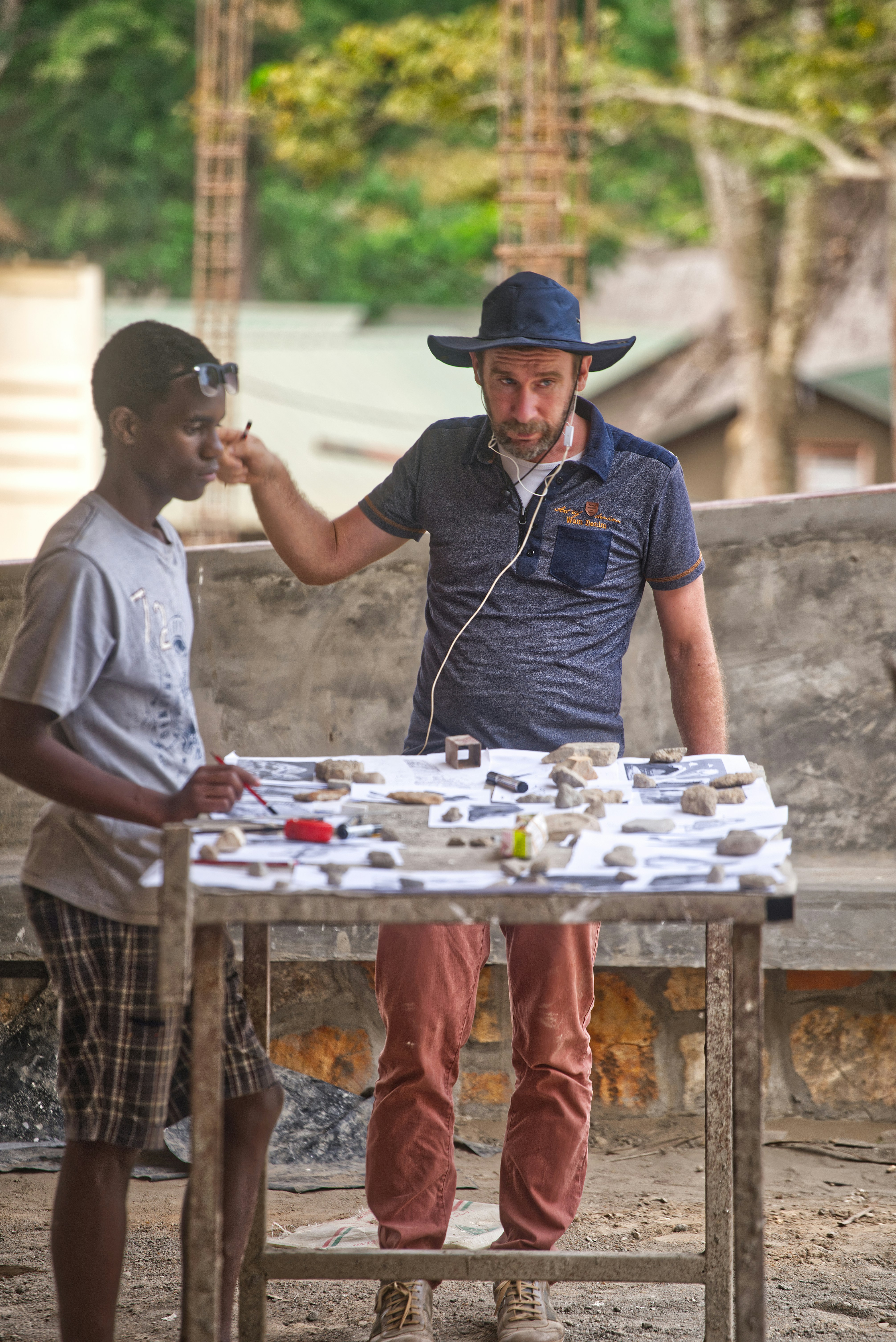 Hands and tools on the rock: a man in a hat guides a roadside stone display