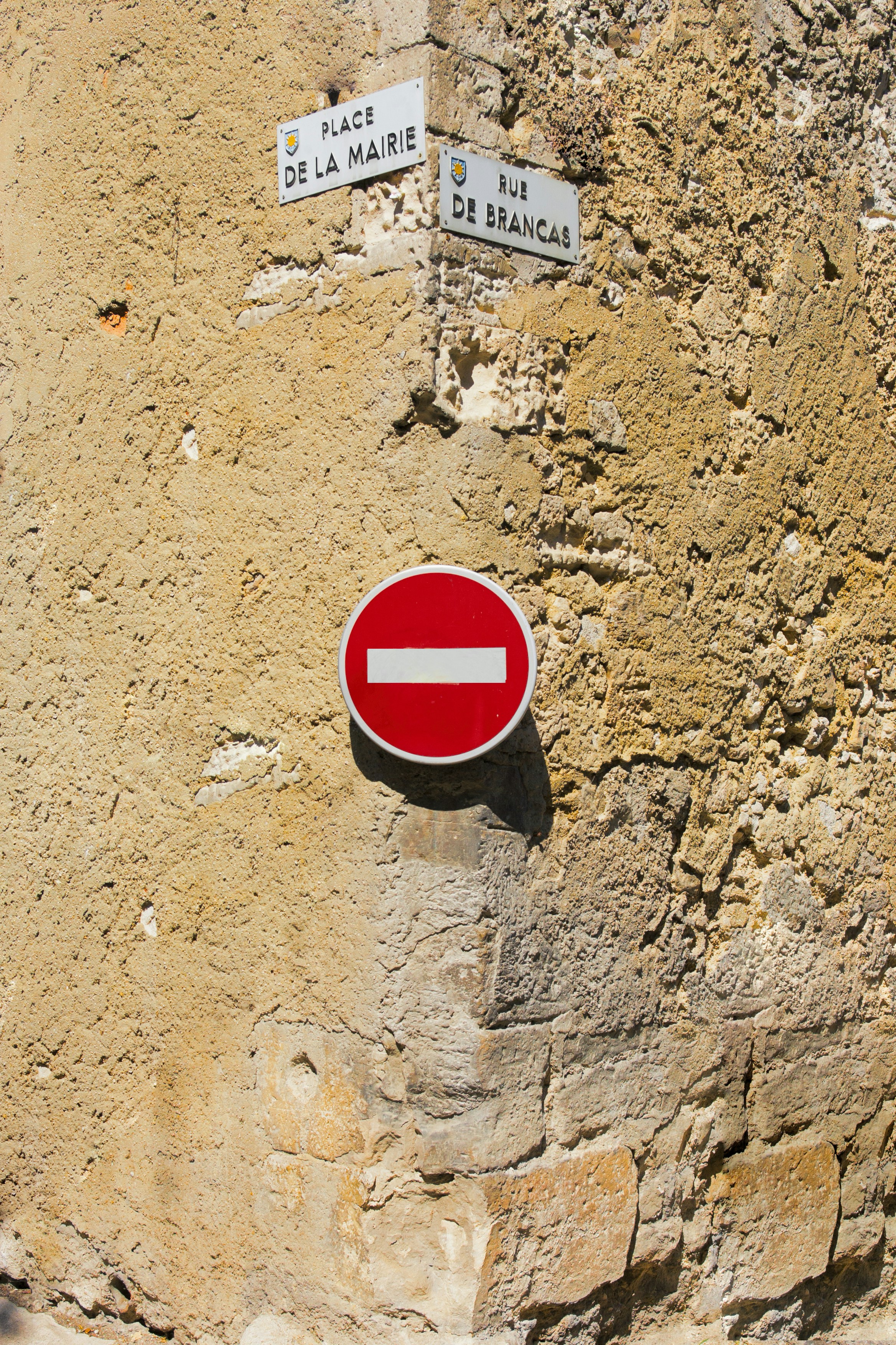 A rustic, textured wall in warm earthy tones features two French street signs for "Place de la Mairie" and "Rue de Brancas", along with a prominent red no-entry sign.