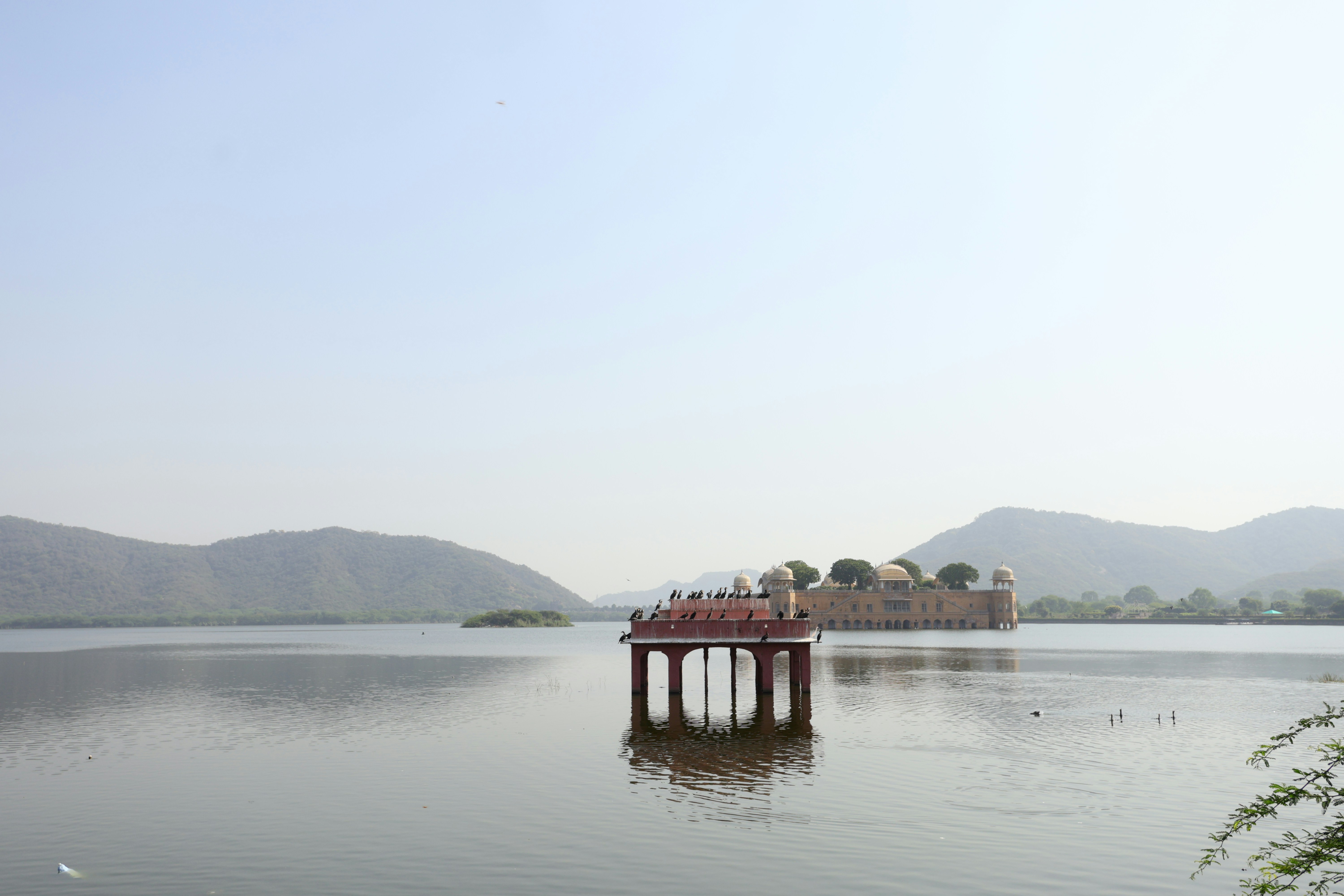 A front view of Jal Mahal with a small arching structure in front of it. The palace is surrounded with water all around with a backdrop of mountain range Aravali.