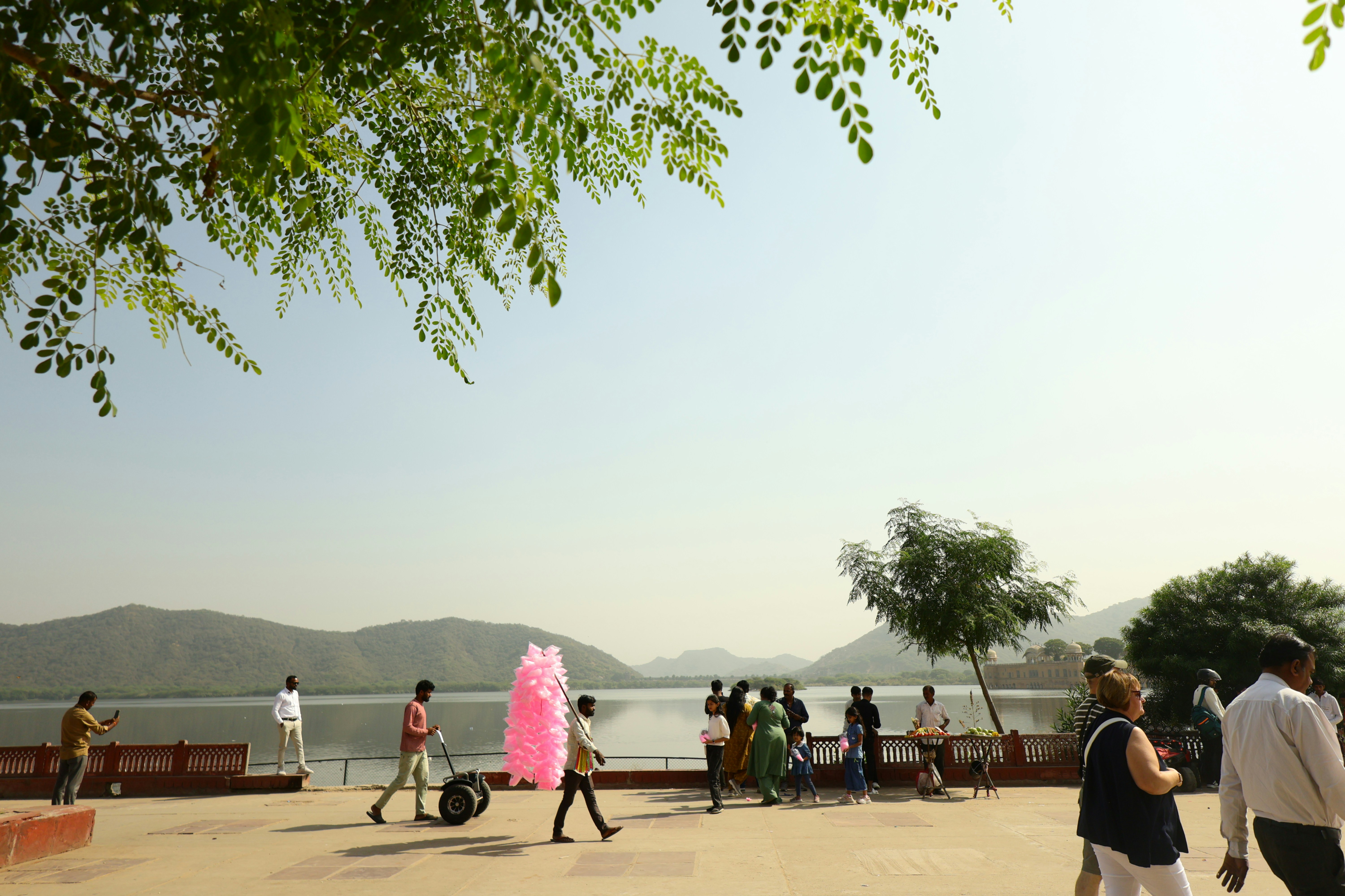 Causal view of people walking on the side of Jal Mahal lake. Focus point is a man selling bright pink colored cotton candy.