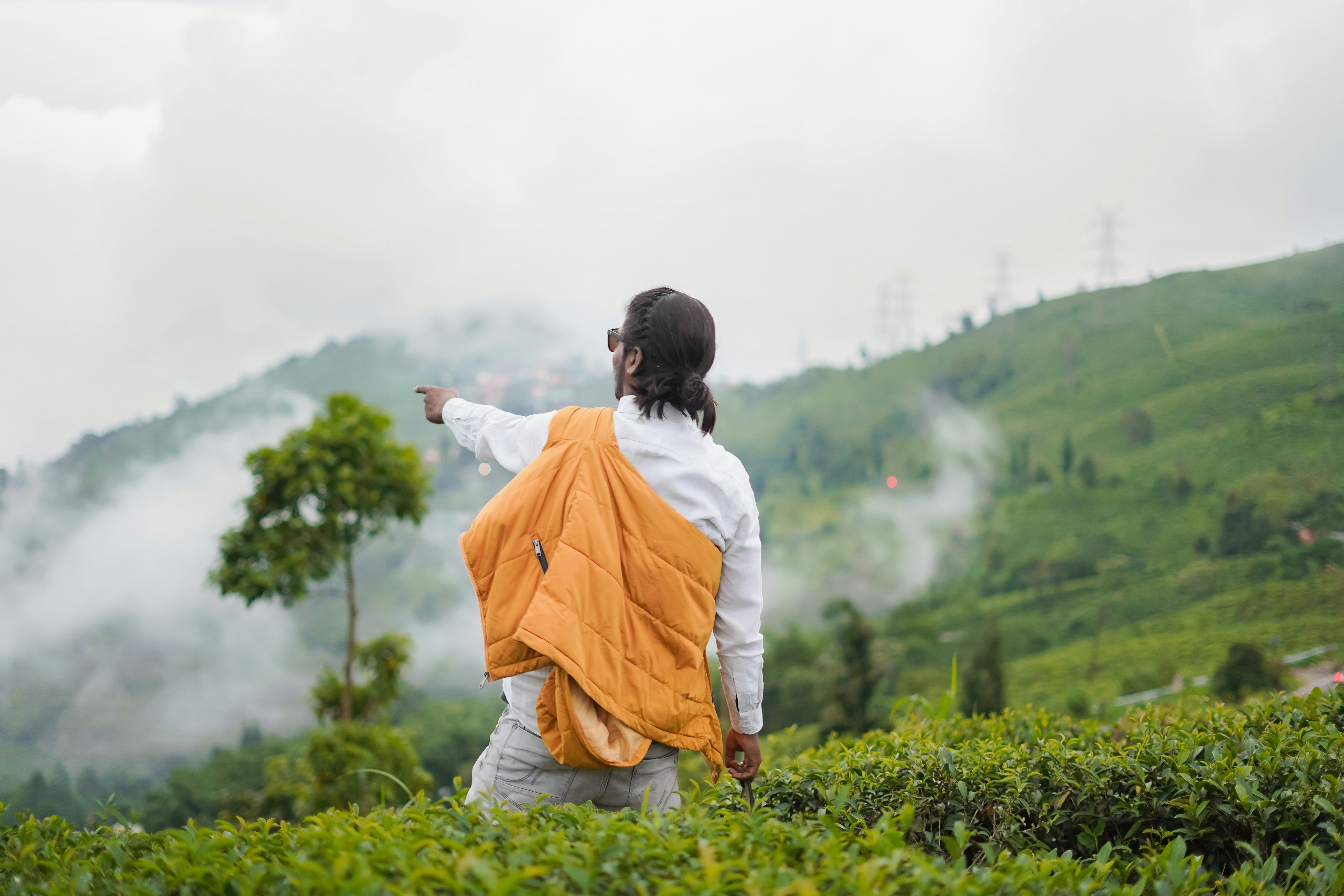 A young man, dressed in a casual grey shirt, stands with his arms wide open, either facing away from the camera in awe of the mountainous view or turning to smile at the camera. The foreground is dominated by vibrant green tea bushes, contrasting with the soft, cloud-covered hills and distant village lights in the background. The mood is one of peace, freedom, and deep connection with nature.