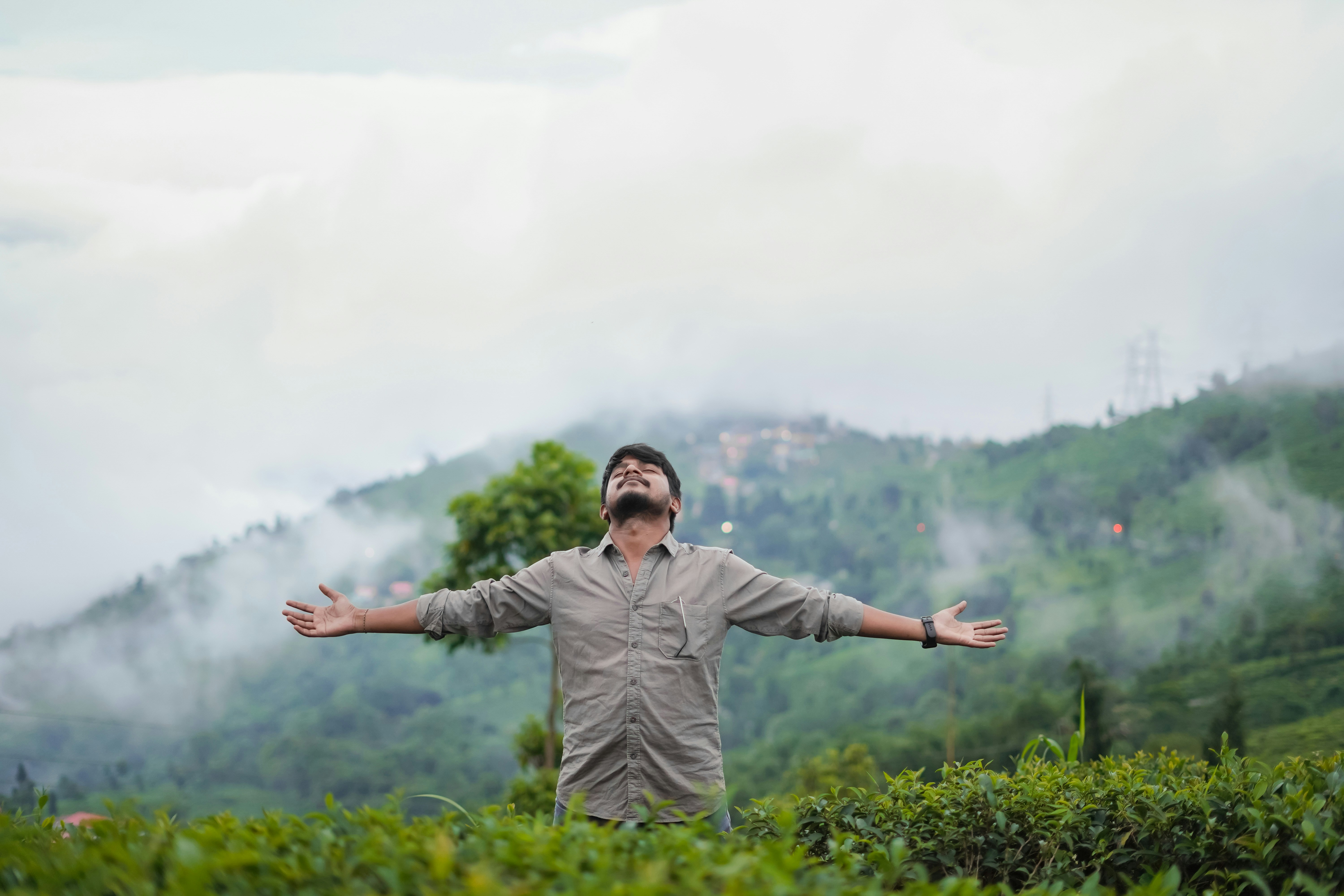 A young man, dressed in a casual grey shirt, stands with his arms wide open, either facing away from the camera in awe of the mountainous view or turning to smile at the camera. The foreground is dominated by vibrant green tea bushes, contrasting with the soft, cloud-covered hills and distant village lights in the background. The mood is one of peace, freedom, and deep connection with nature.