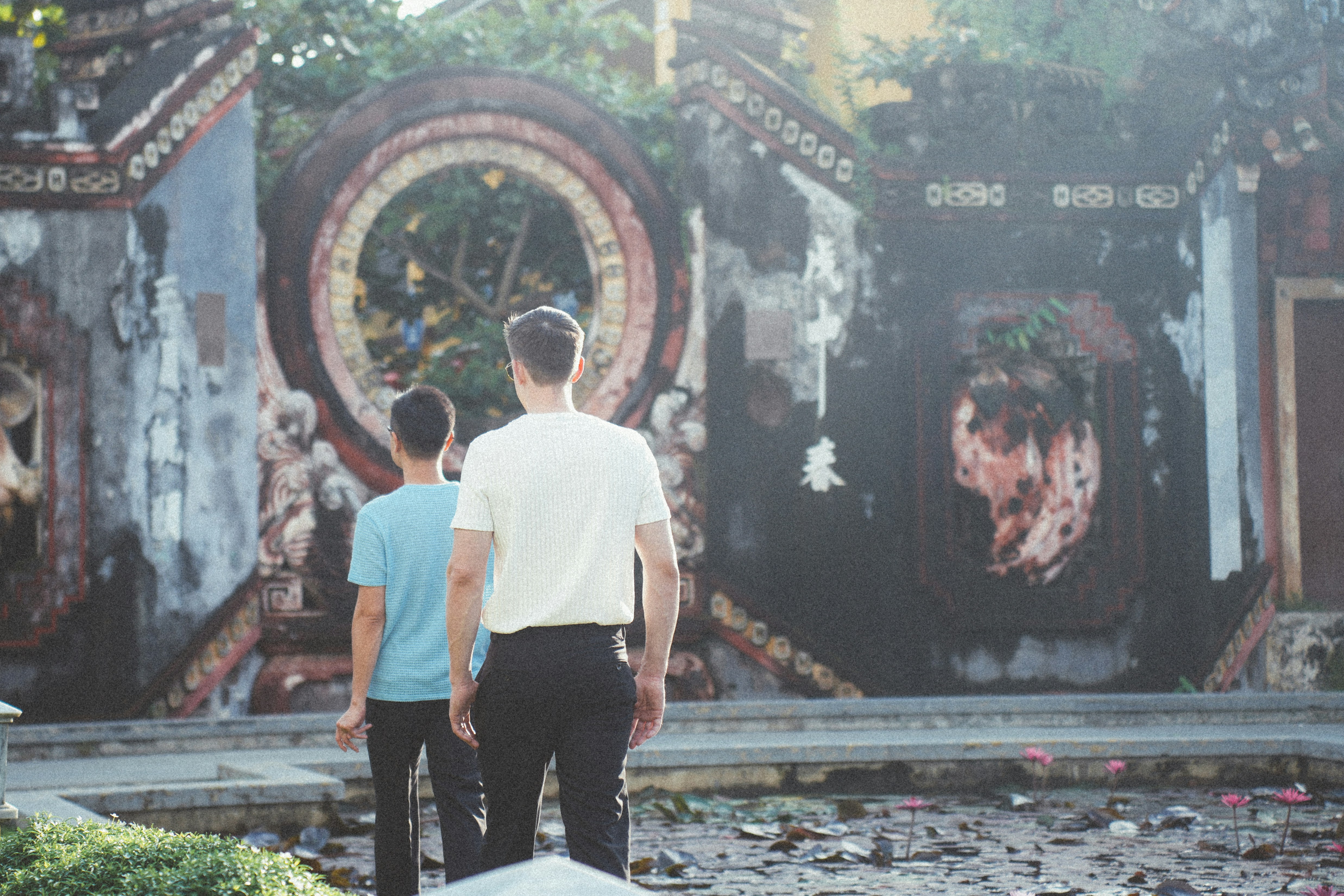 Two Visitors Confront the Circular Shrine Gate in a Weathered Temple Courtyard