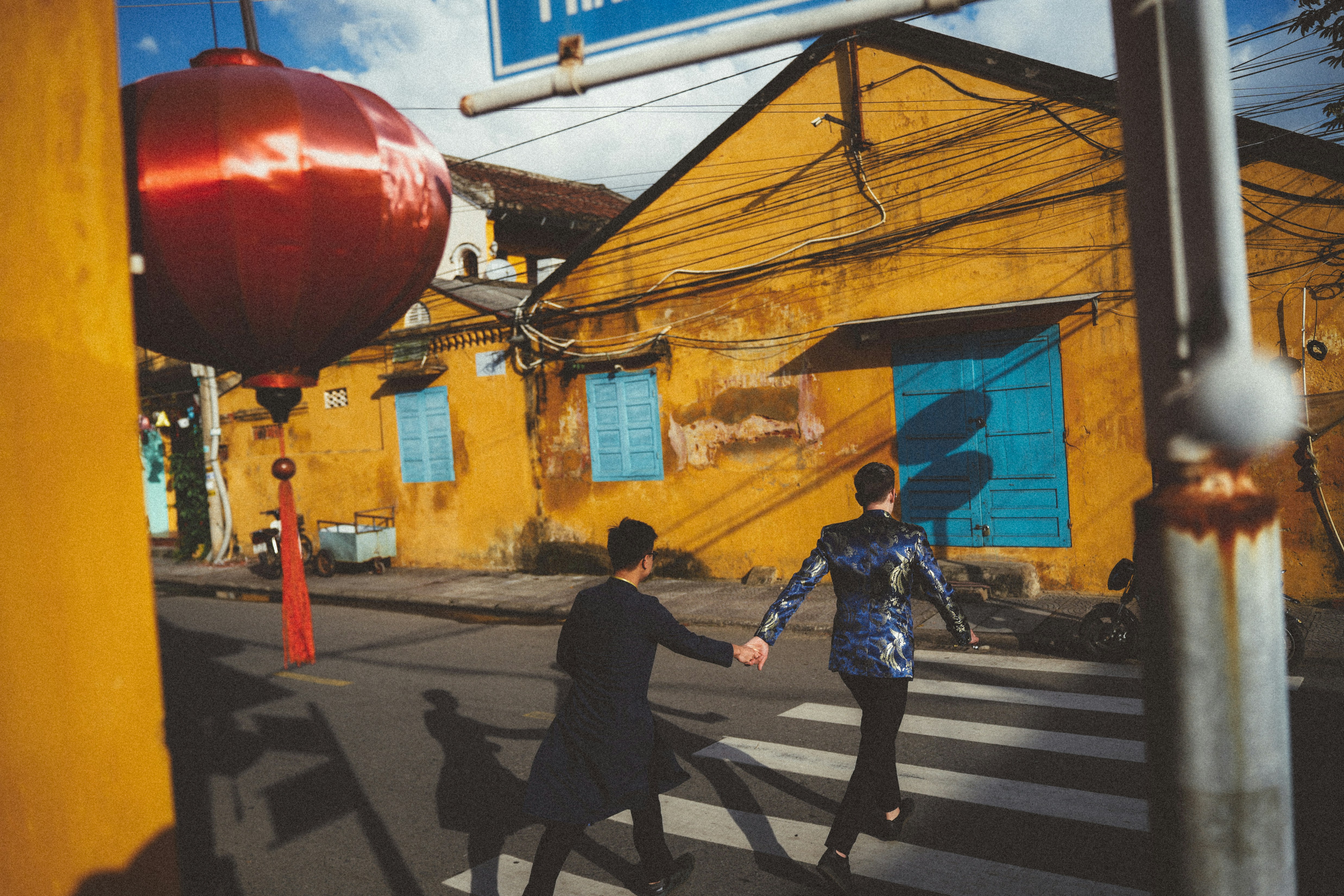 Two pedestrians cross under a sunlit yellow facade as a red lantern sways above