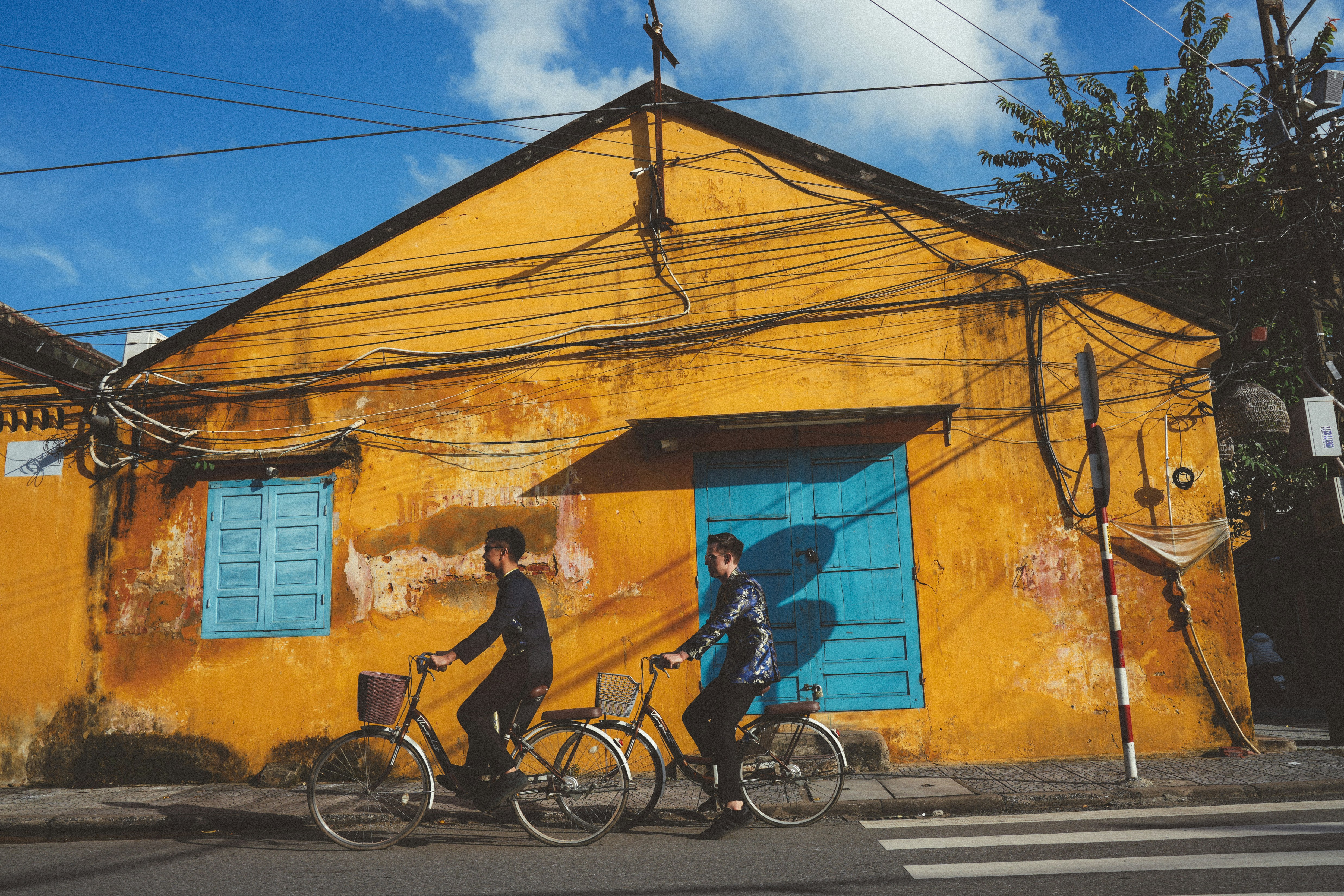 Two cyclists glide past a sunlit ochre storefront with indigo doors