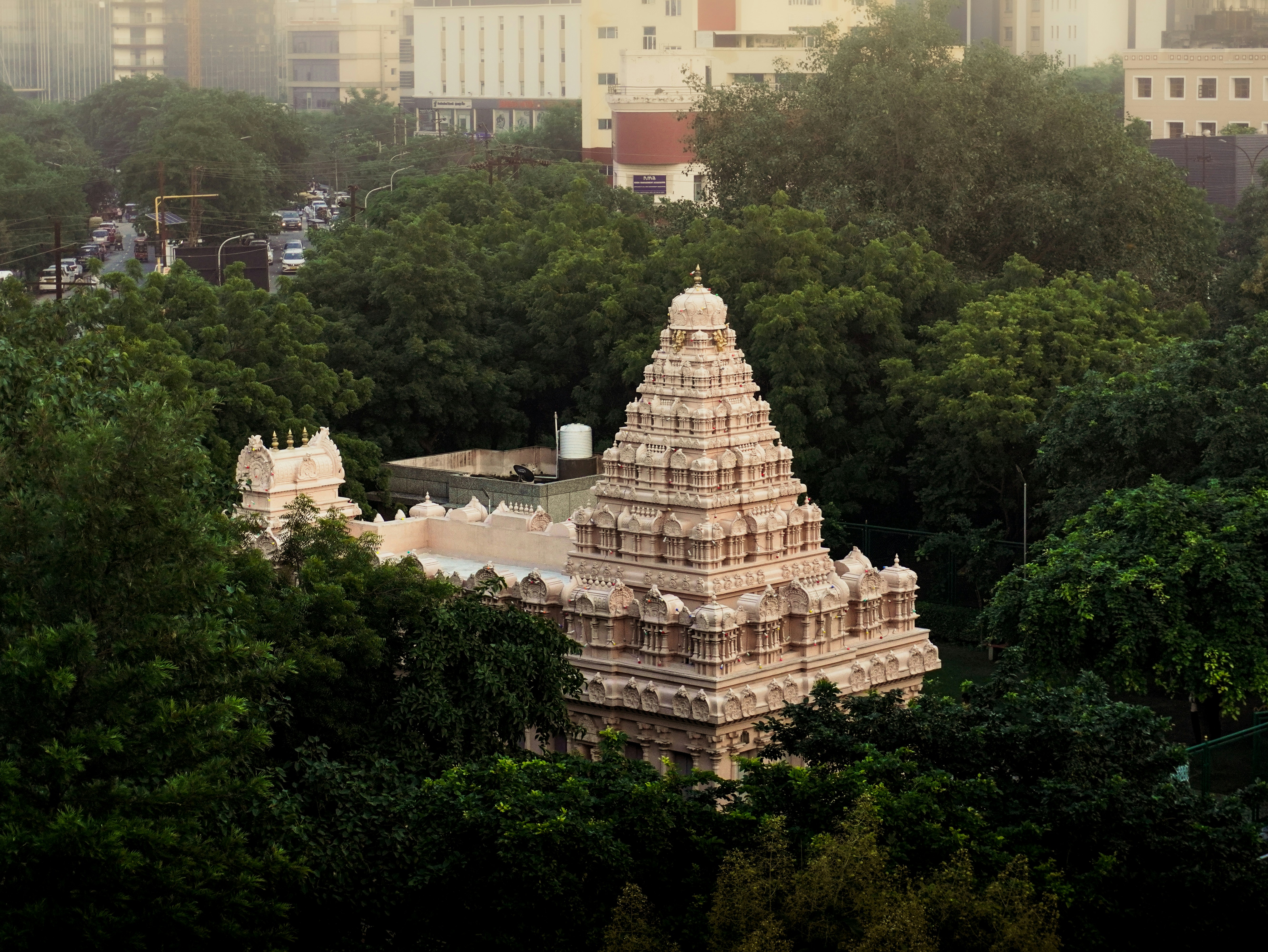 A beautifully detailed temple emerges from a dense canopy of lush green trees, its pale stone architecture glowing softly in the warm evening light. Surrounded by the bustle of an urban skyline, the temple stands as a serene contrast—an oasis of history, spirituality, and intricate craftsmanship nestled within the modern cityscape. The elevated perspective highlights both the temple’s grandeur and its harmonious blend with nature.