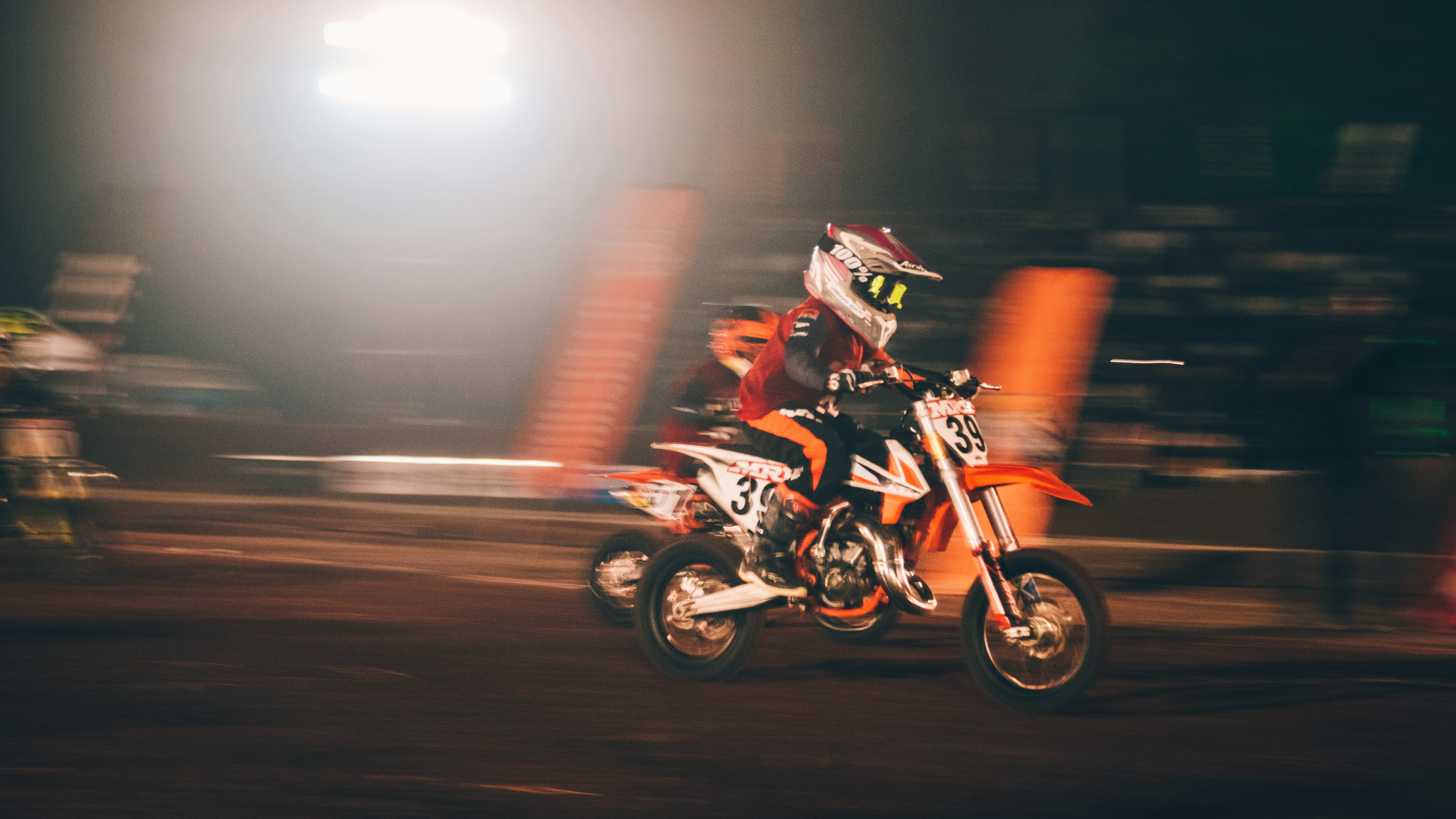 A motocross rider wearing a red and black racing suit and a patterned helmet speeds along the track on an orange and white bike displaying the number 39. Motion blur in the background emphasizes the high speed, with hints of other racers and trackside elements visible. Bright overhead lighting fades into darkness, creating a dramatic and energetic racing atmosphere.