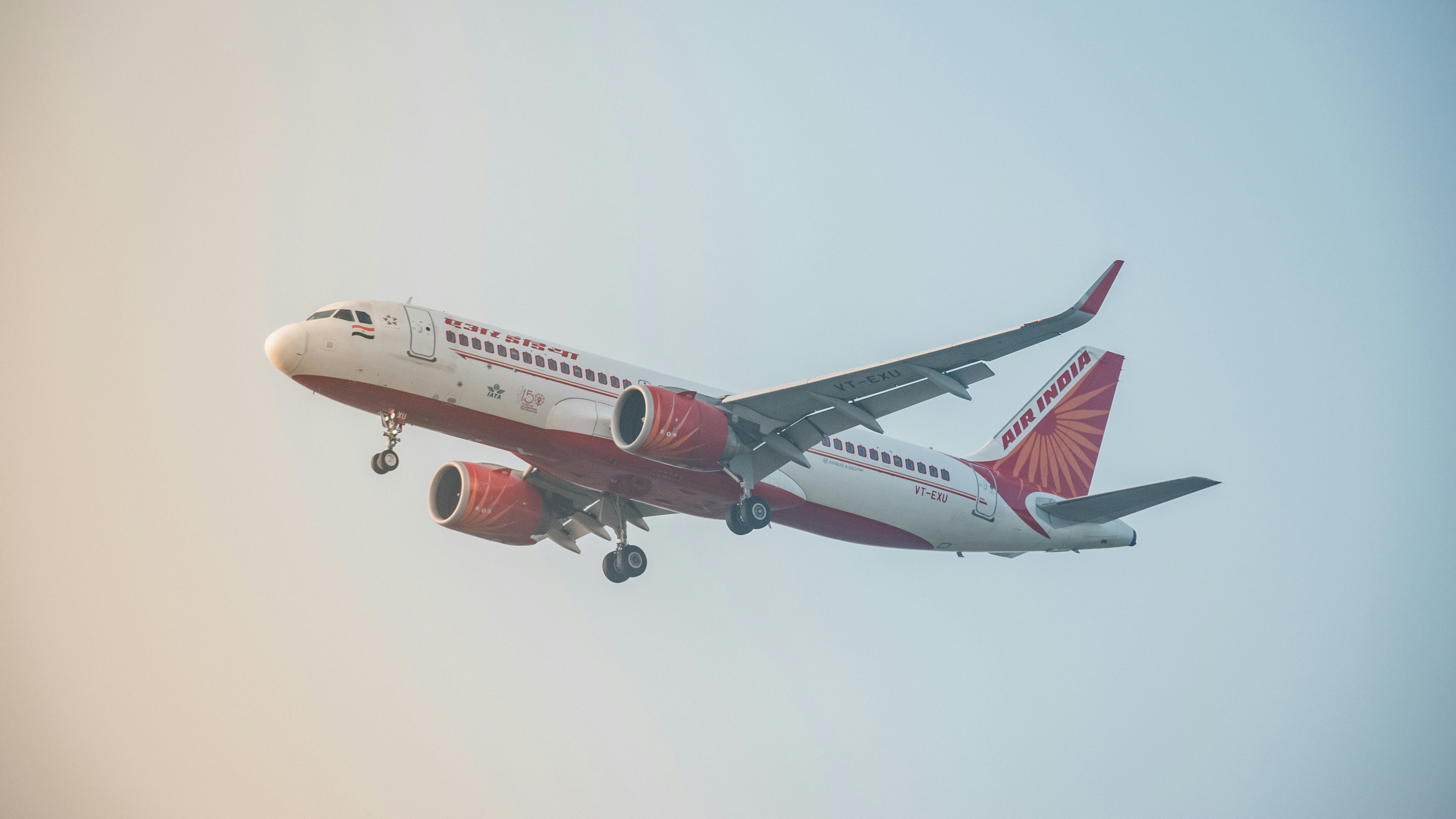 An Air India aircraft with landing gear down flies against a gradient sky transitioning from warm tones to pale blue. The plane’s white body features a red stripe, red engine accents, and the Air India sunburst logo on the tail. The registration number VT-EXU is visible as the aircraft angles slightly, suggesting takeoff or landing.