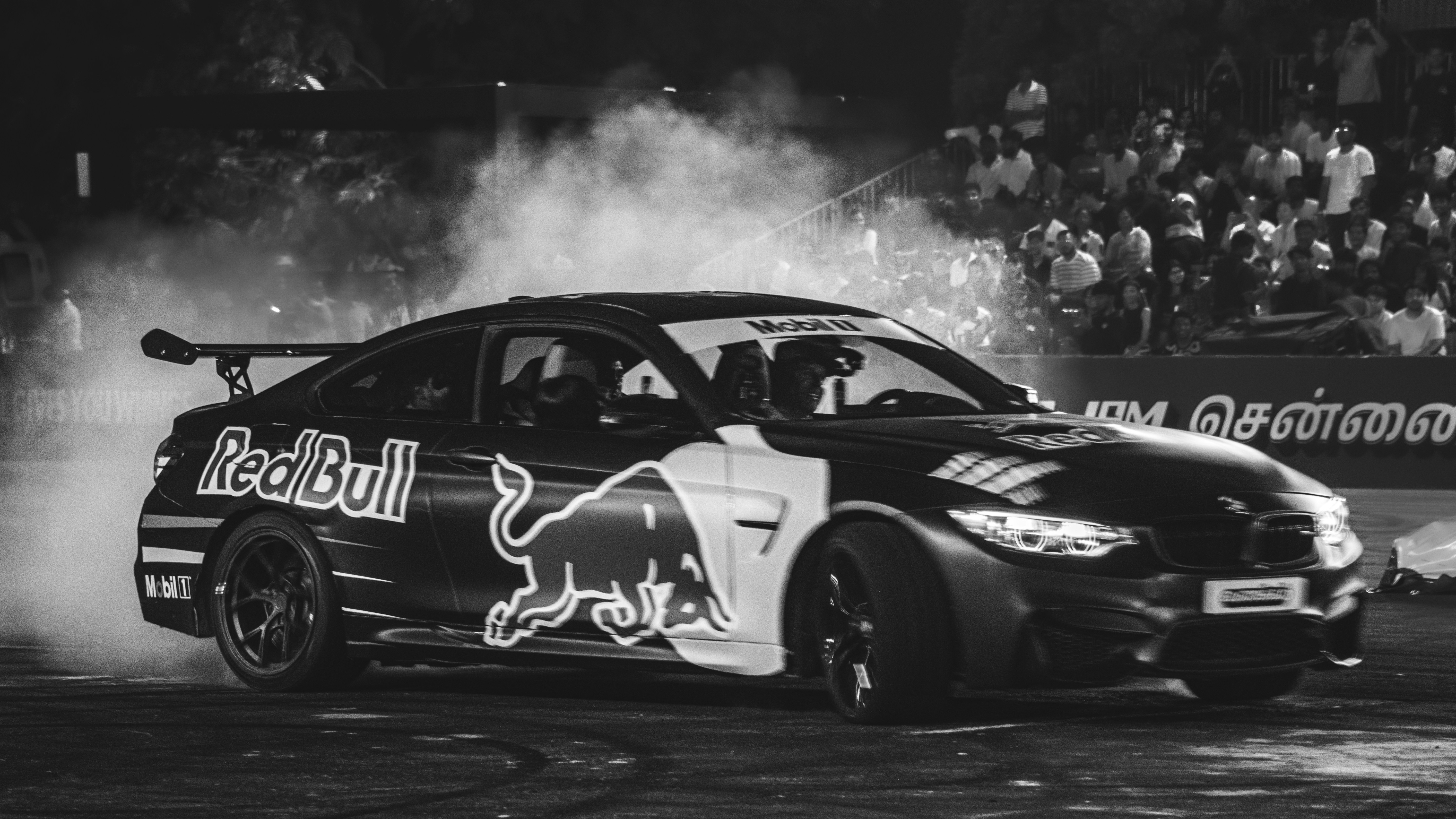 A black and white photograph of a Red Bull–branded drift car sliding across the track, producing a dense cloud of smoke from the rear tires. A large crowd watches from the stands in the background as the car drifts with precision, its headlights cutting through the smoky atmosphere.