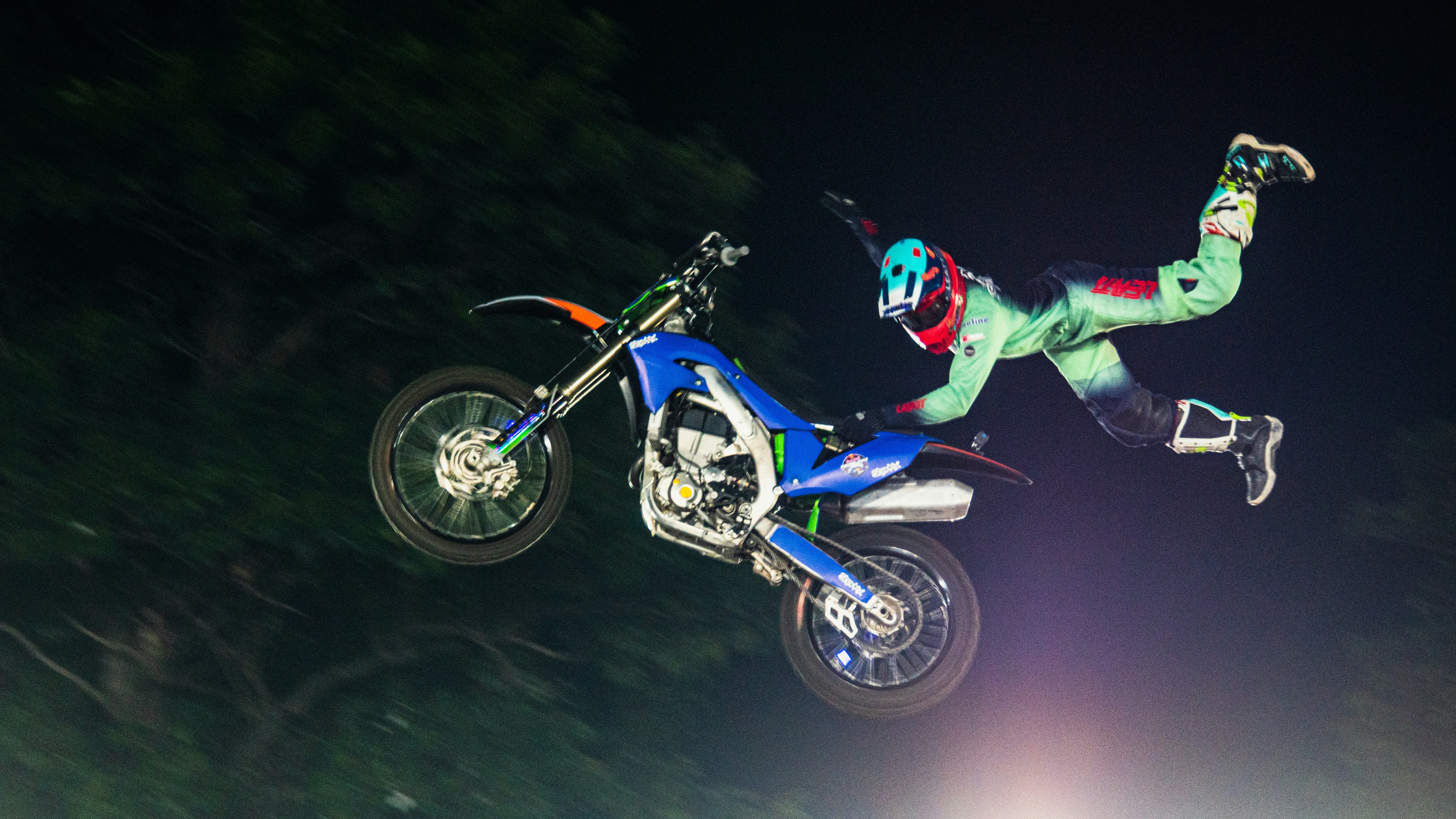 A freestyle motocross rider performs an airborne trick at night, extending their body off the side of a blue dirt bike. The bike and rider are suspended high in the air against a dark background, emphasizing motion and athletic skill.