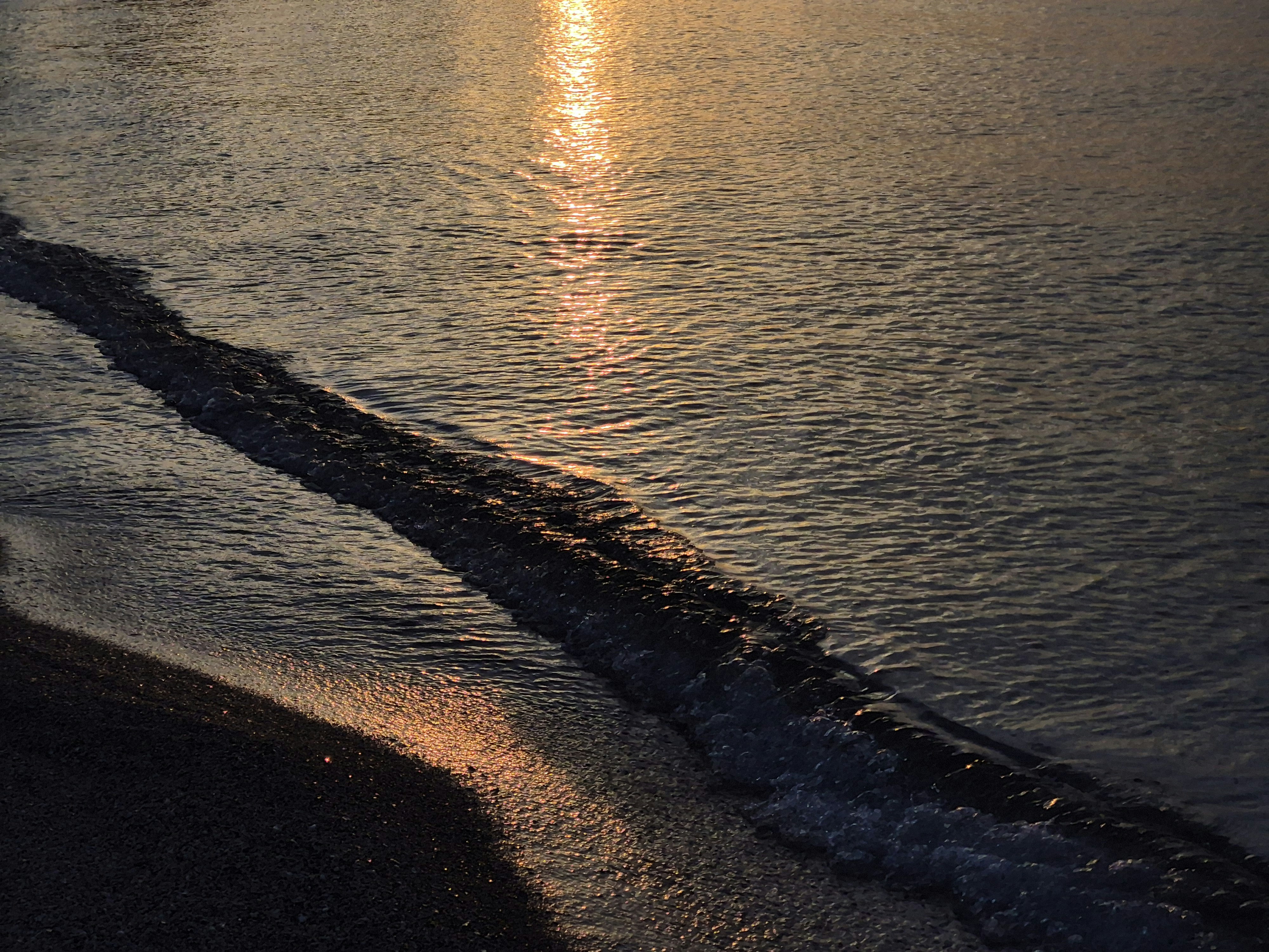 Gentle waves lap a dark shore at sunset.