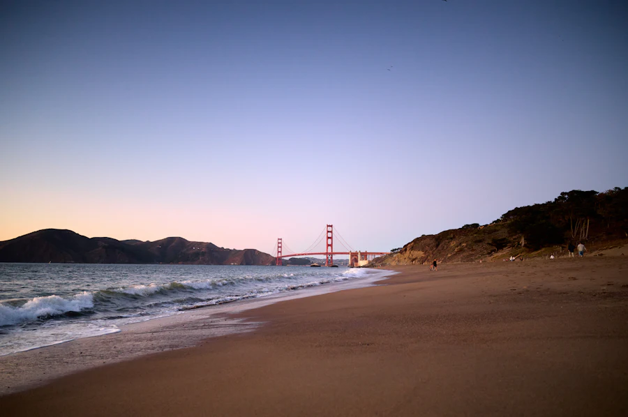 Sunlit shoreline with gentle waves and a distant bridge silhouette, conveying serene coastal atmosphere.