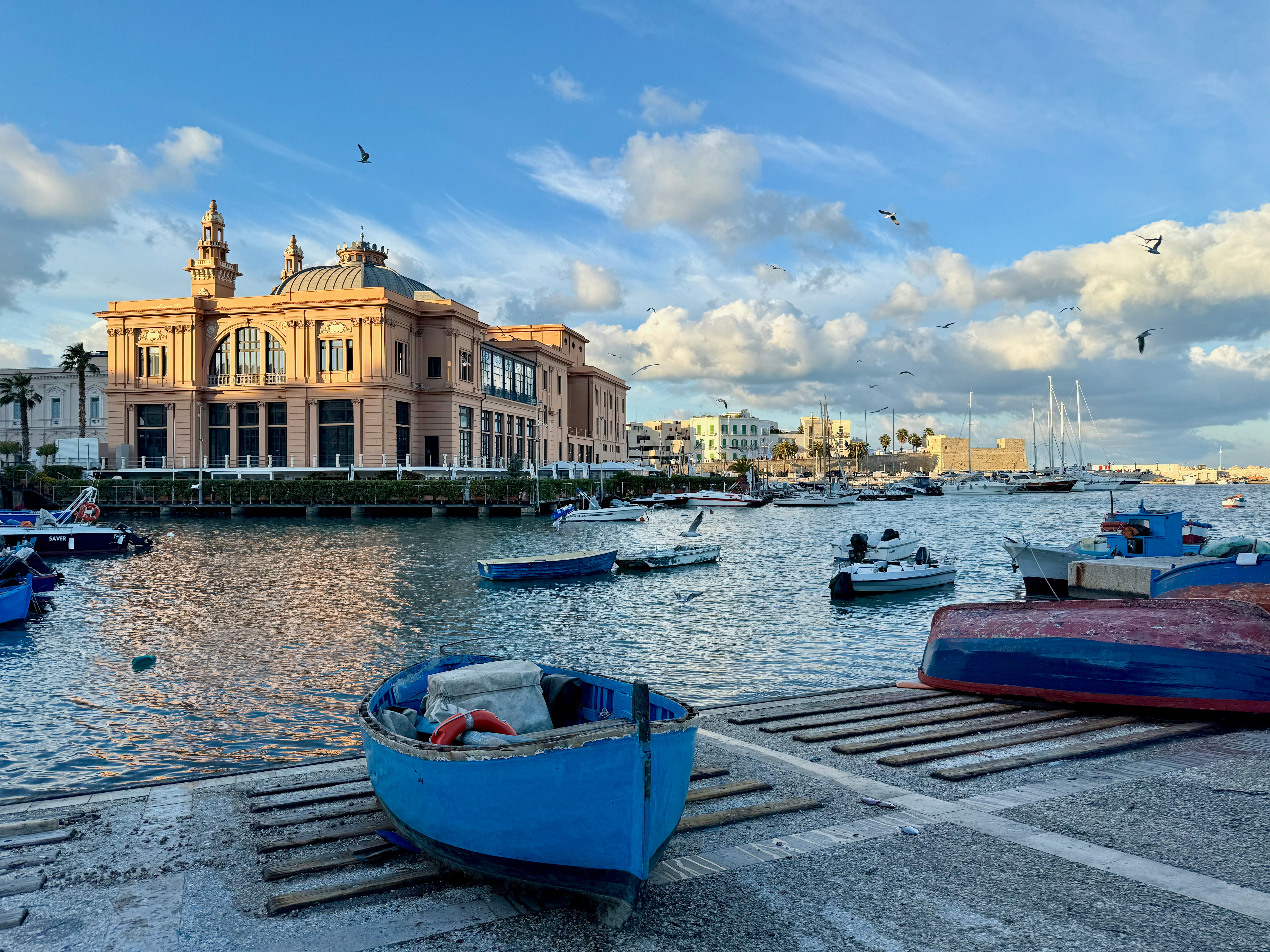 Colorful boats float in the harbor of Bari, Italy with a stately waterfront building and dramatic clouds illuminated by the evening sun.