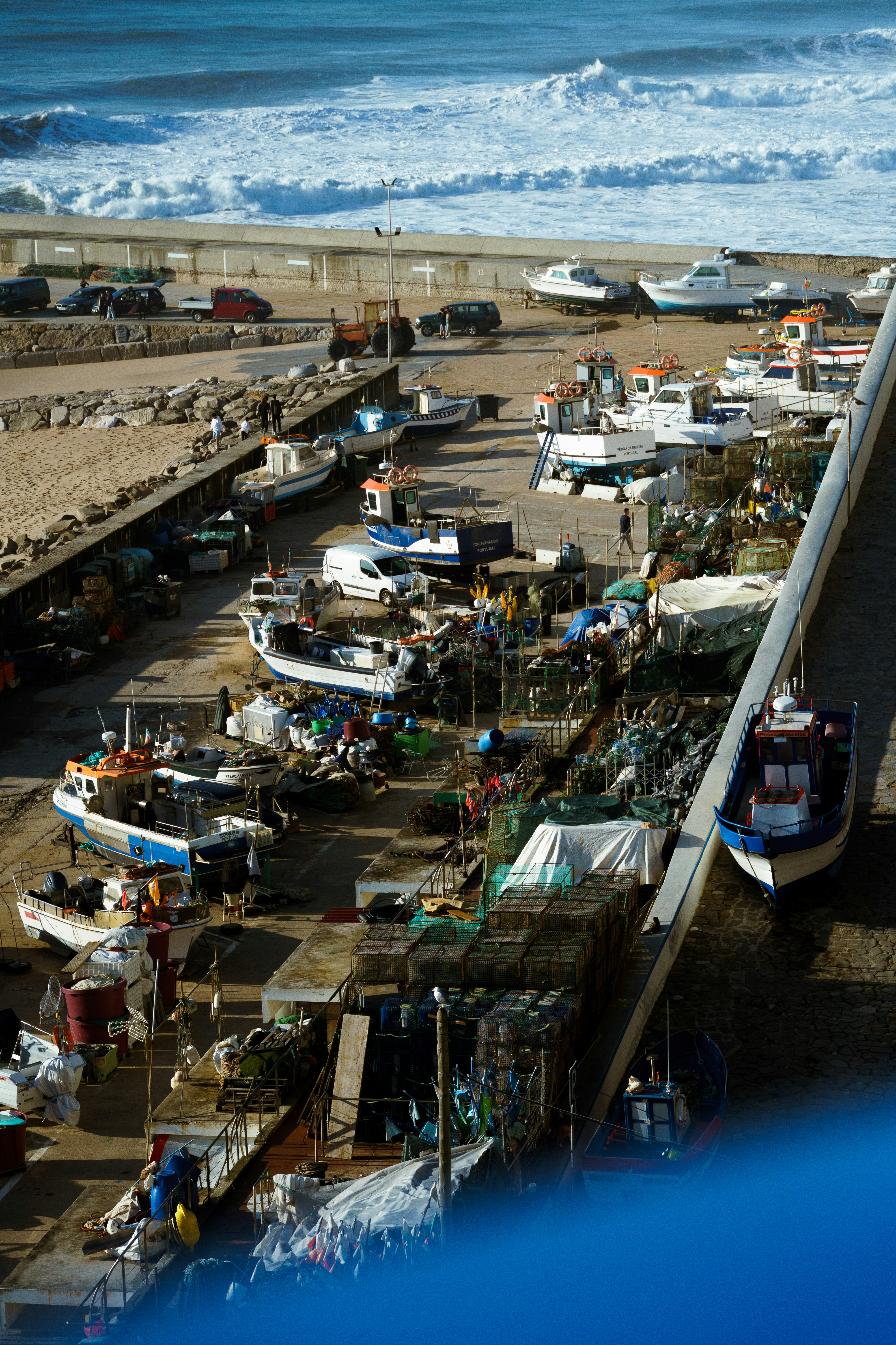 Small fishing boats resting in a coastal harbor while strong ocean waves crash against the breakwater, capturing the contrast between calm harbor life and the raw power of the sea.