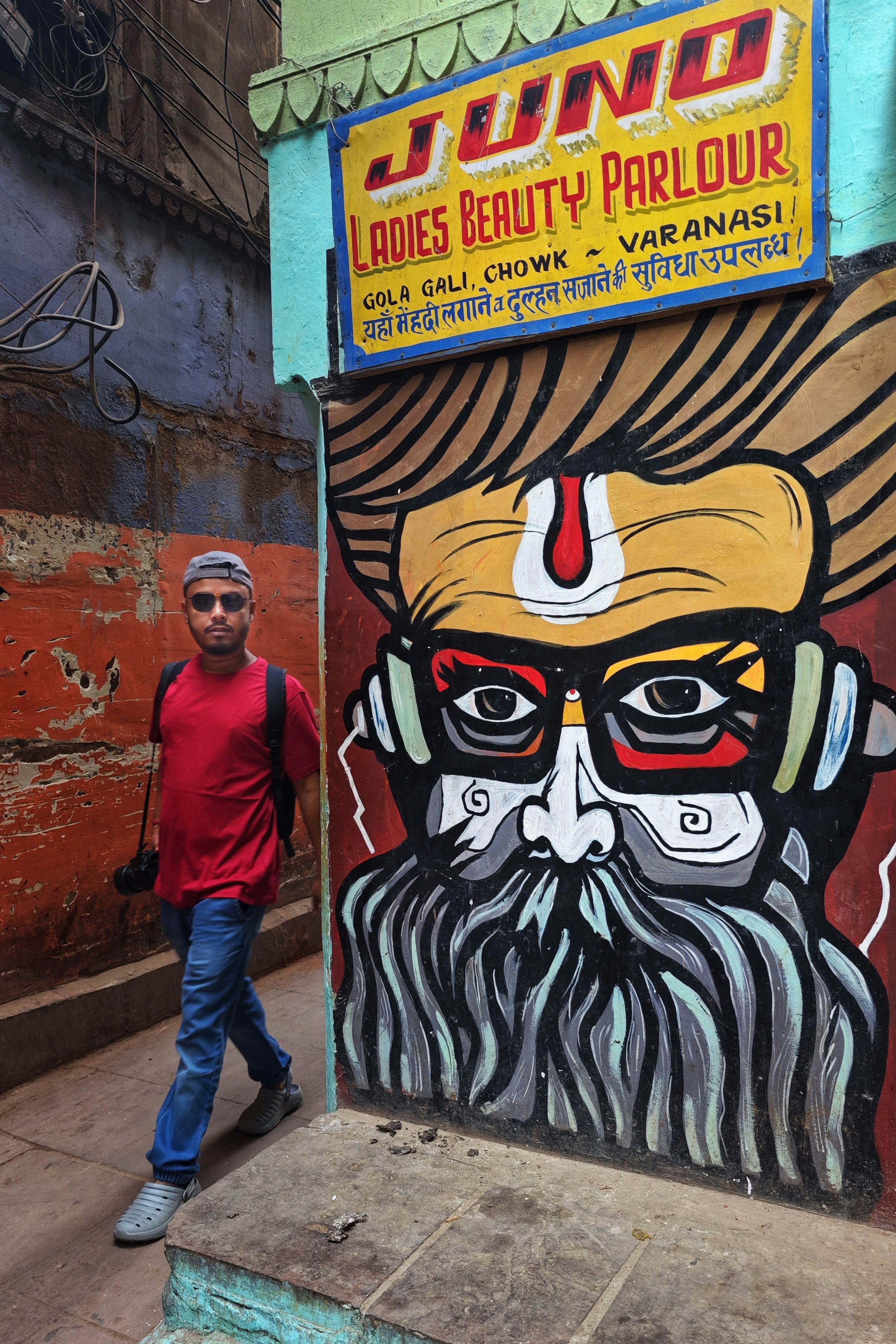 Red- shirted wanderer beside a vibrant mural of a masked guru