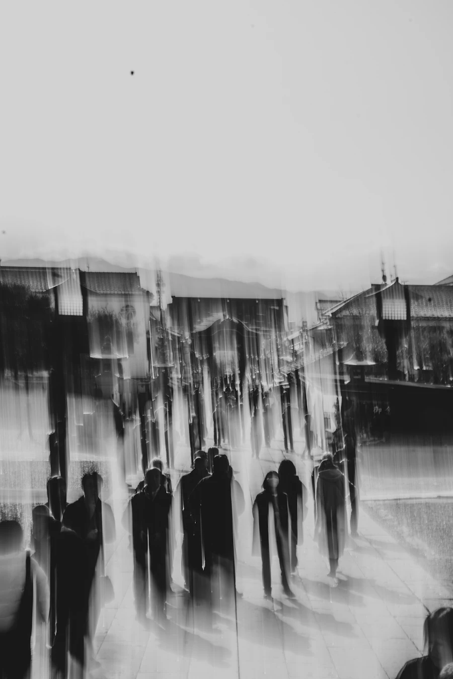 Monochrome composition of blurred, overlapping silhouettes in a narrow urban corridor, suggesting movement and repetition through glass or reflection. The scene evokes a moody, abstract street environment with ghostlike figures.