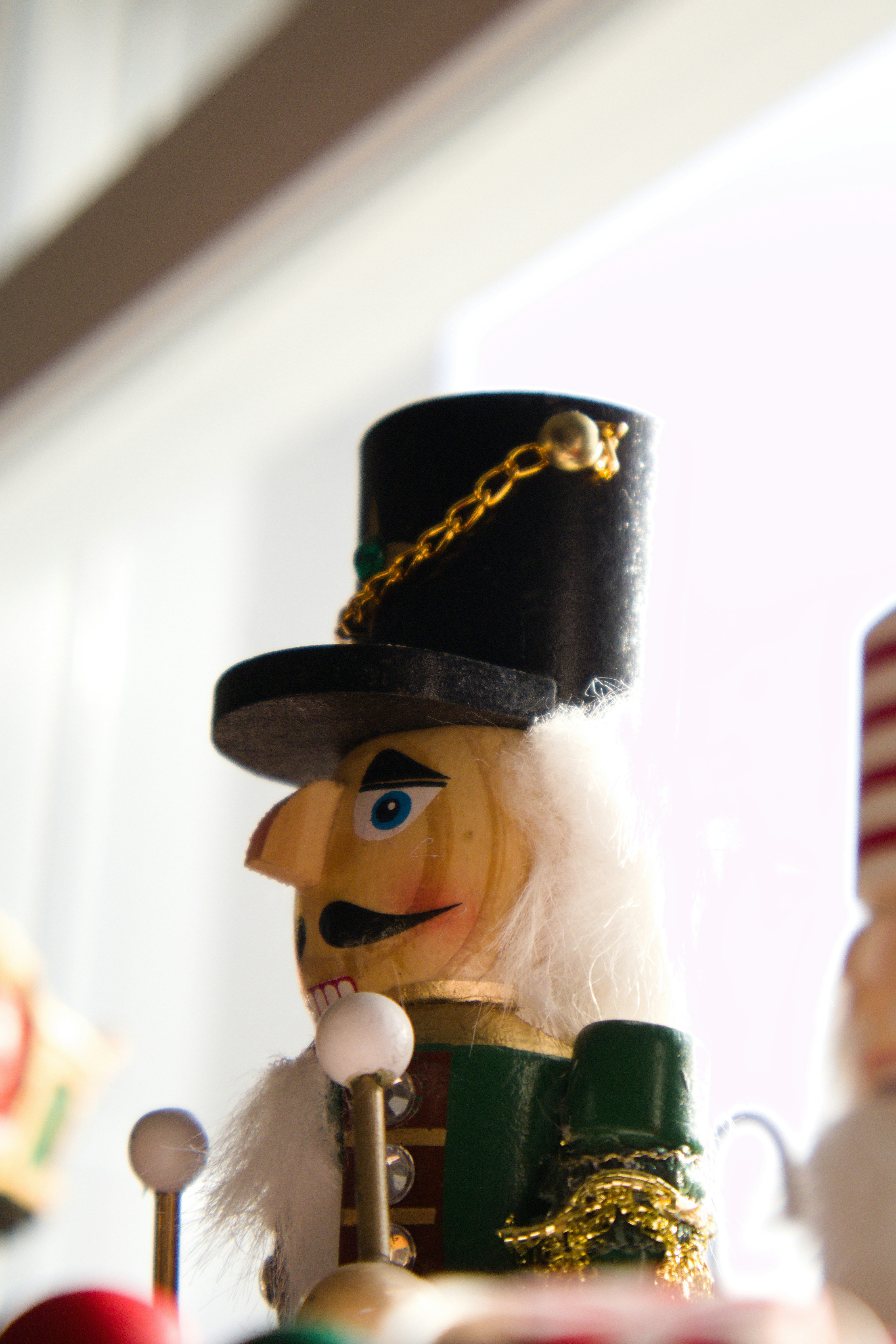 Close-up of a wooden nutcracker figure with a black hat and white beard, spotlighted among blurred props in a bright display, a logo-like decorative toy scene.