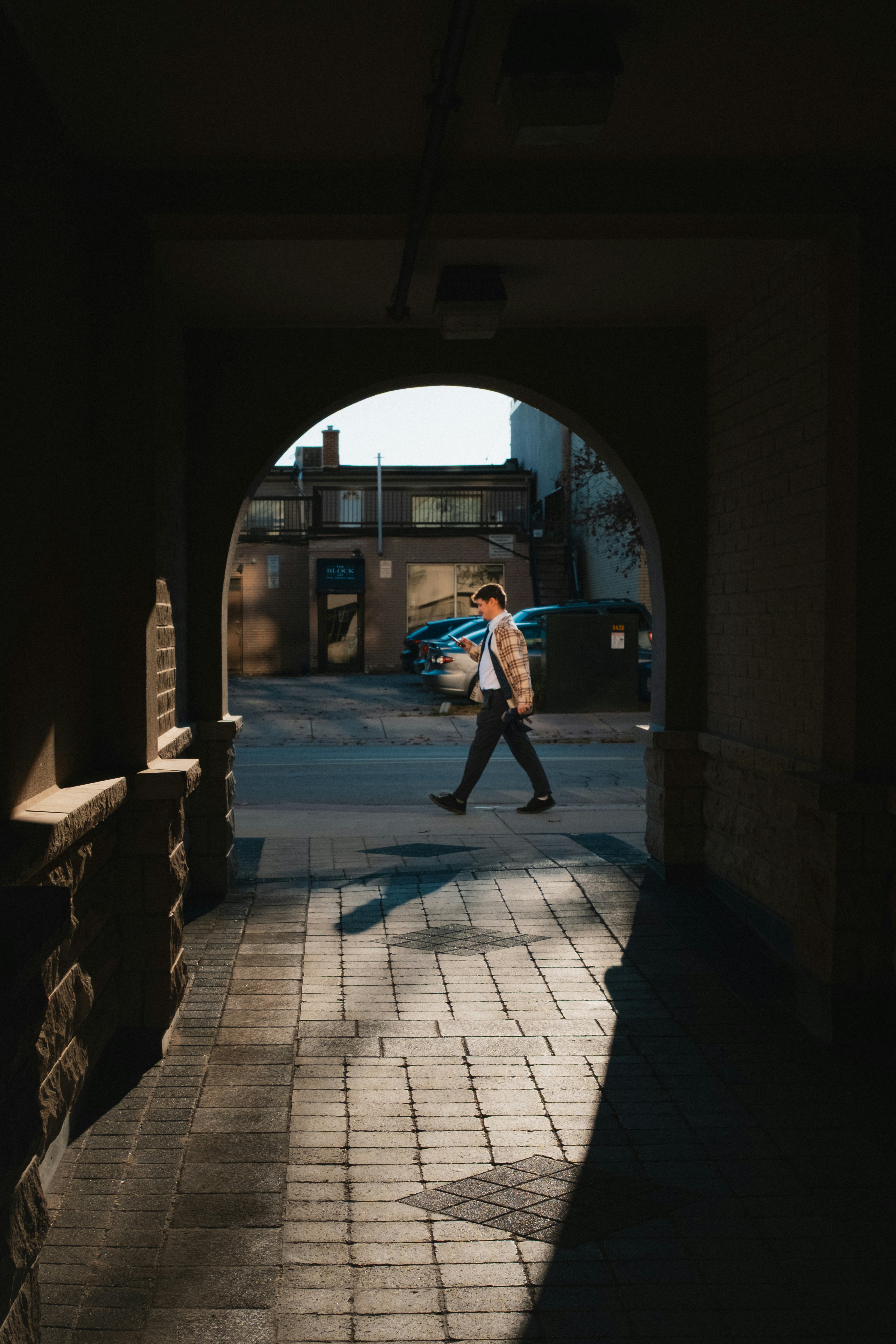 A man walks through an archway, caught in a hard slice of late-afternoon light. Deep shadows, brick and paving stones frame the scene with a clean, cinematic street-photography feel.
