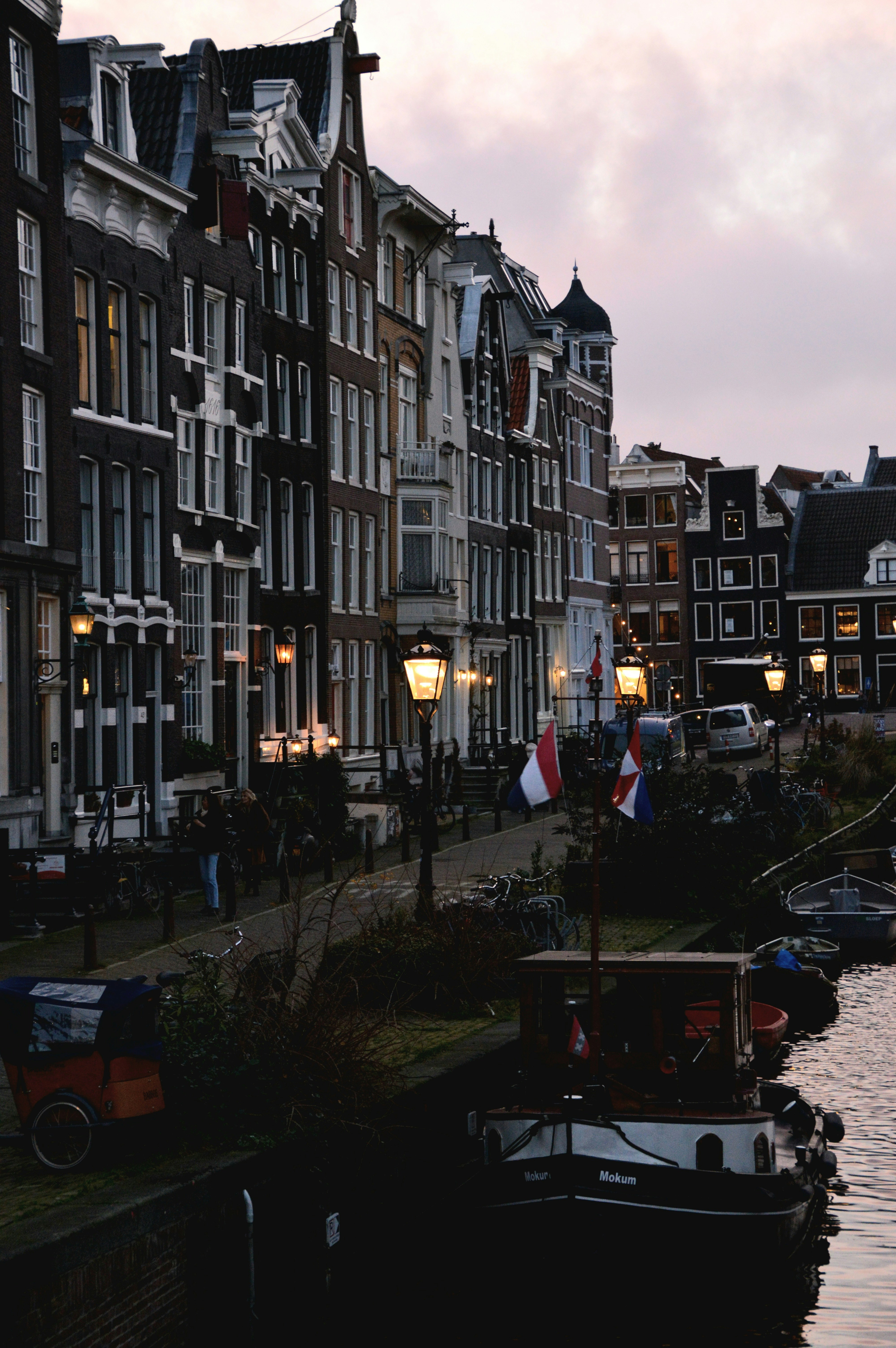 A quiet evening along one of Amsterdam’s historic canals, where narrow Dutch houses line the water and warm streetlights reflect softly on the surface. Boats rest along the canal as the city settles into dusk, capturing the timeless atmosphere of Amsterdam just after sunset.