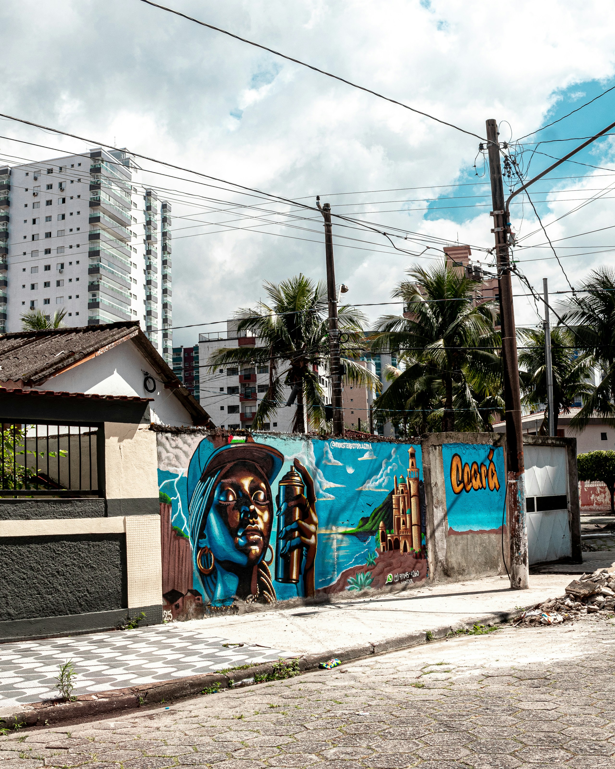 A vibrant mural depicting a woman holding a spray can adorns a concrete wall in a city. The artwork features a stylized portrait of a woman with closed eyes and a spray can in her hand, set against a backdrop of a tropical landscape with a castle and palm trees. The word "Ceará" is prominently displayed in bold lettering. The mural is located on a street with distinctive patterned sidewalks and a backdrop of modern buildings and palm trees, suggesting an urban tropical setting.