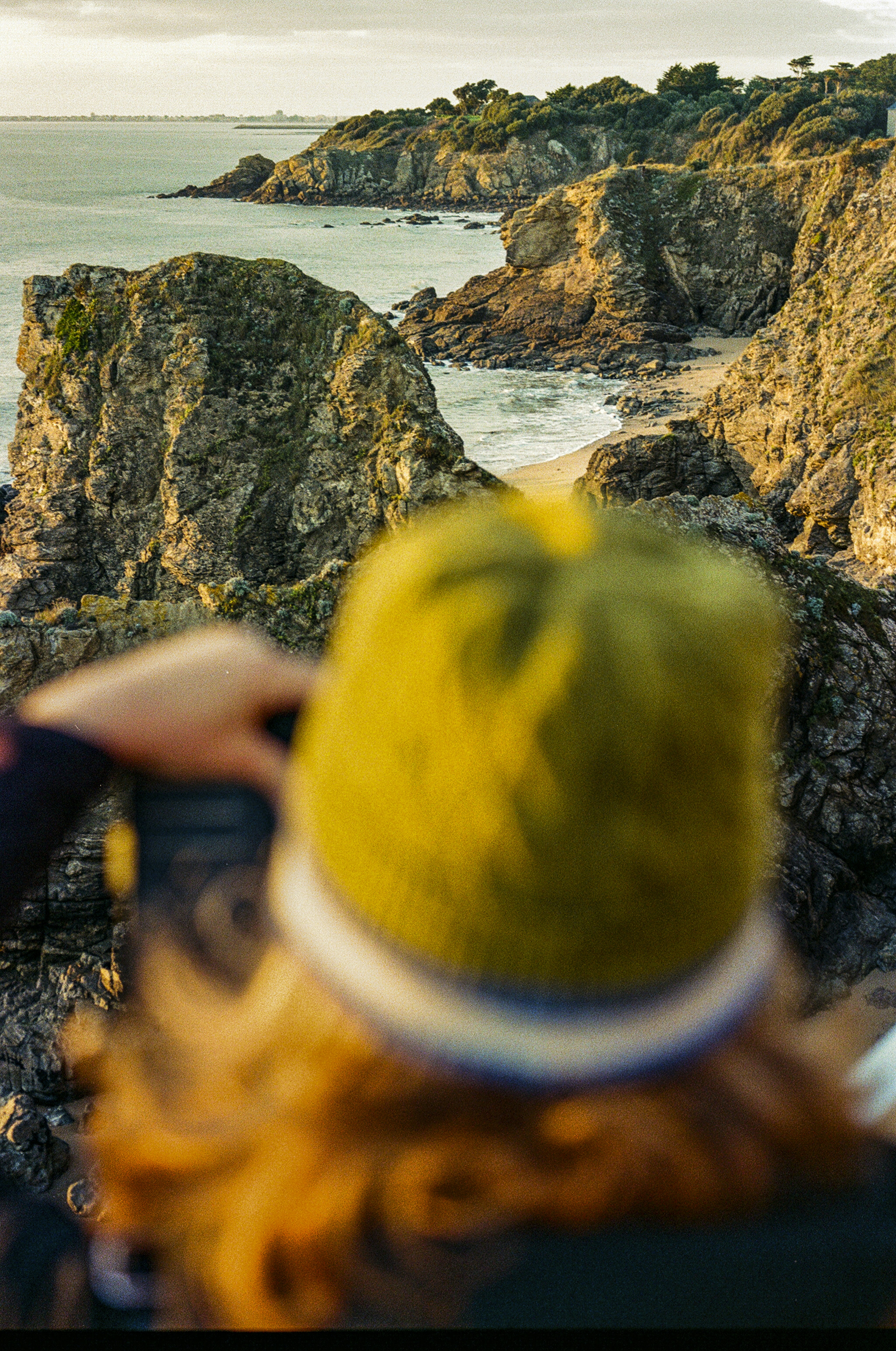 A person wearing a green beanie takes a photograph over a rocky coastline at sunset in Pornichet, France. The foreground is intentionally out of focus, while the cliffs and sea catch the last warm light of the day. Shot on 35mm film with a Nikon F3 HP and a 55mm lens, using Kodak Gold 200, giving the scene a soft grain and natural colors.