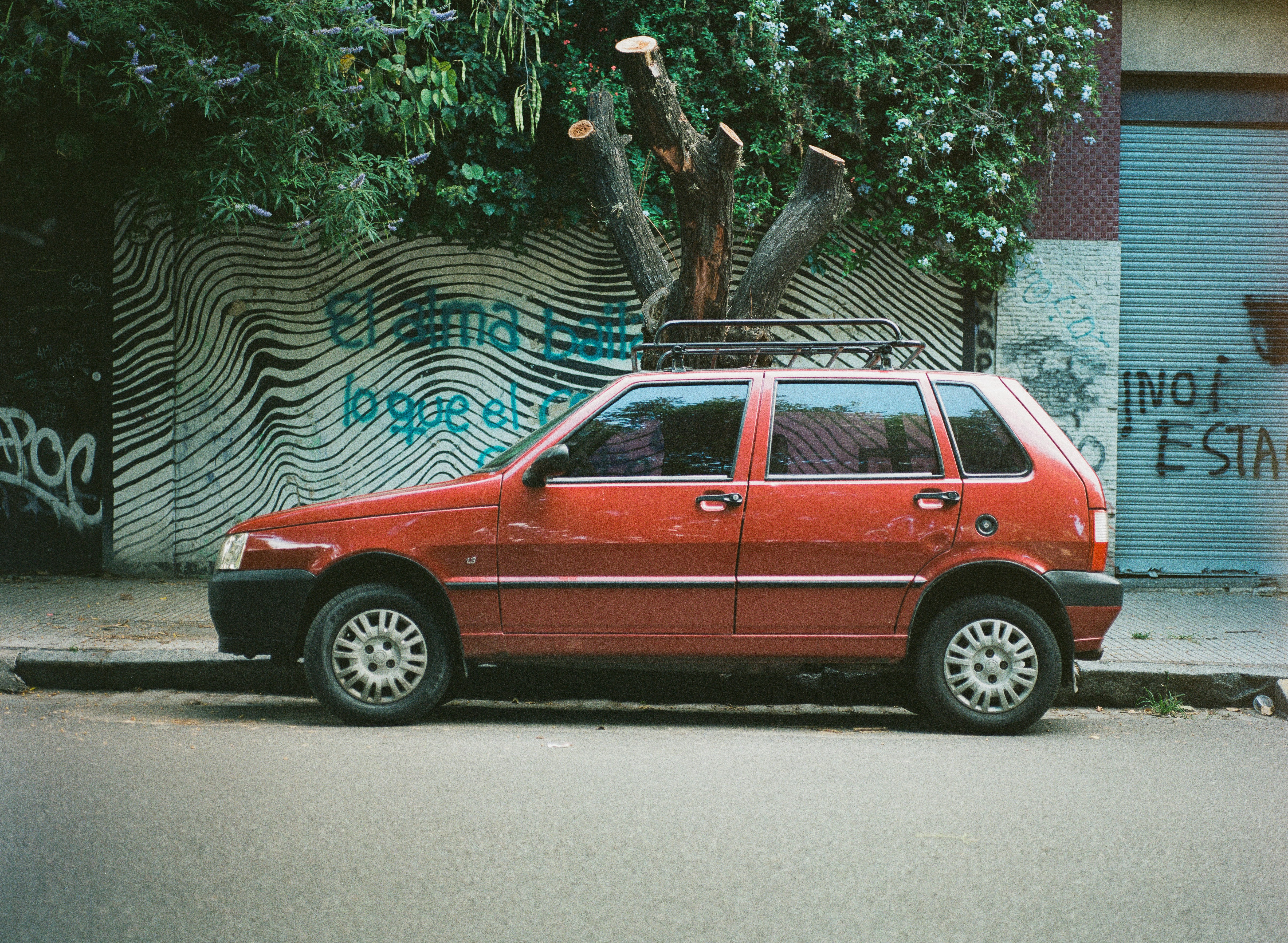 A red car parked along a quiet street with textured walls and greenery in the background, capturing everyday city life.