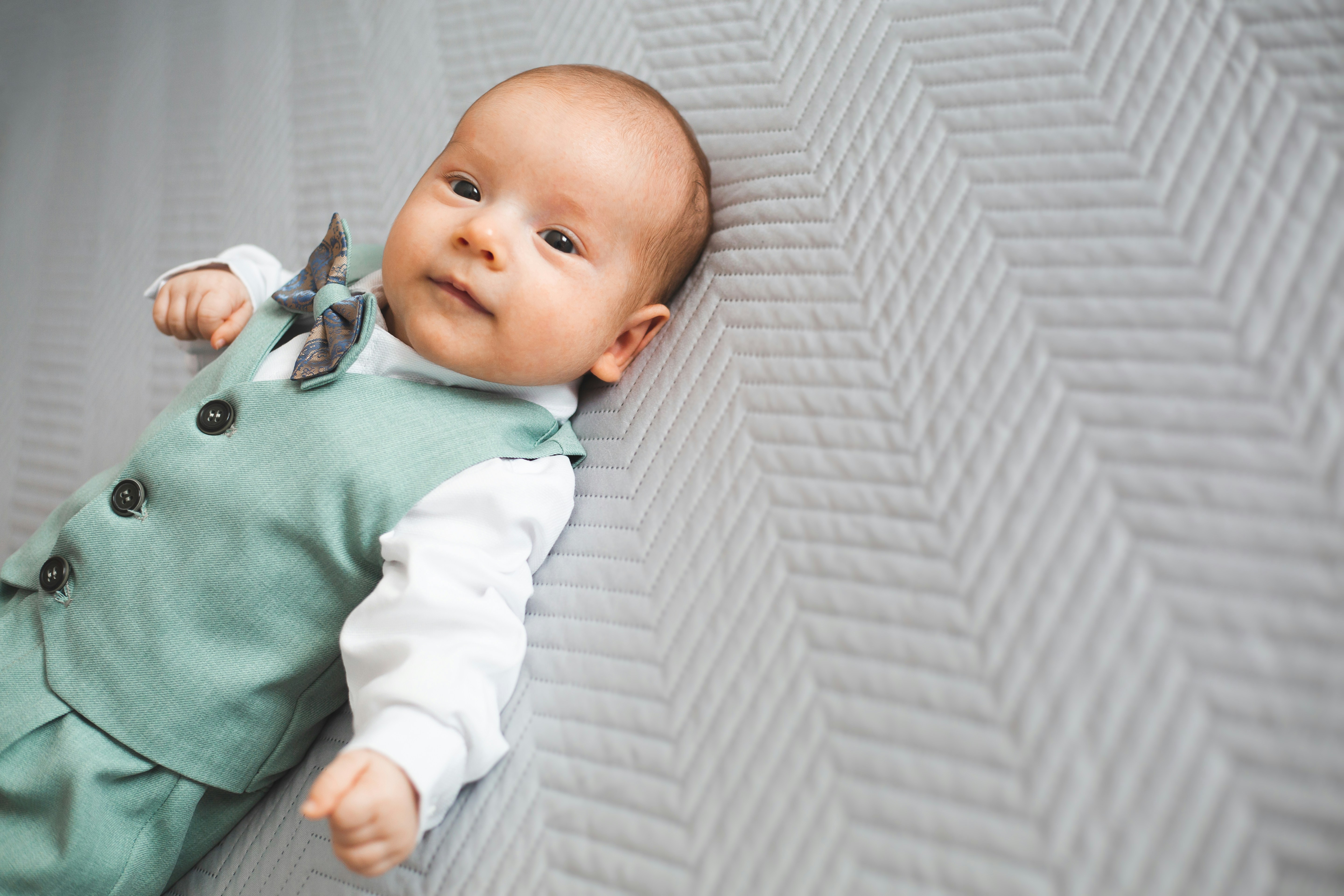 Young baby boy dressed up, lying on his back on bed, looking towards camera.