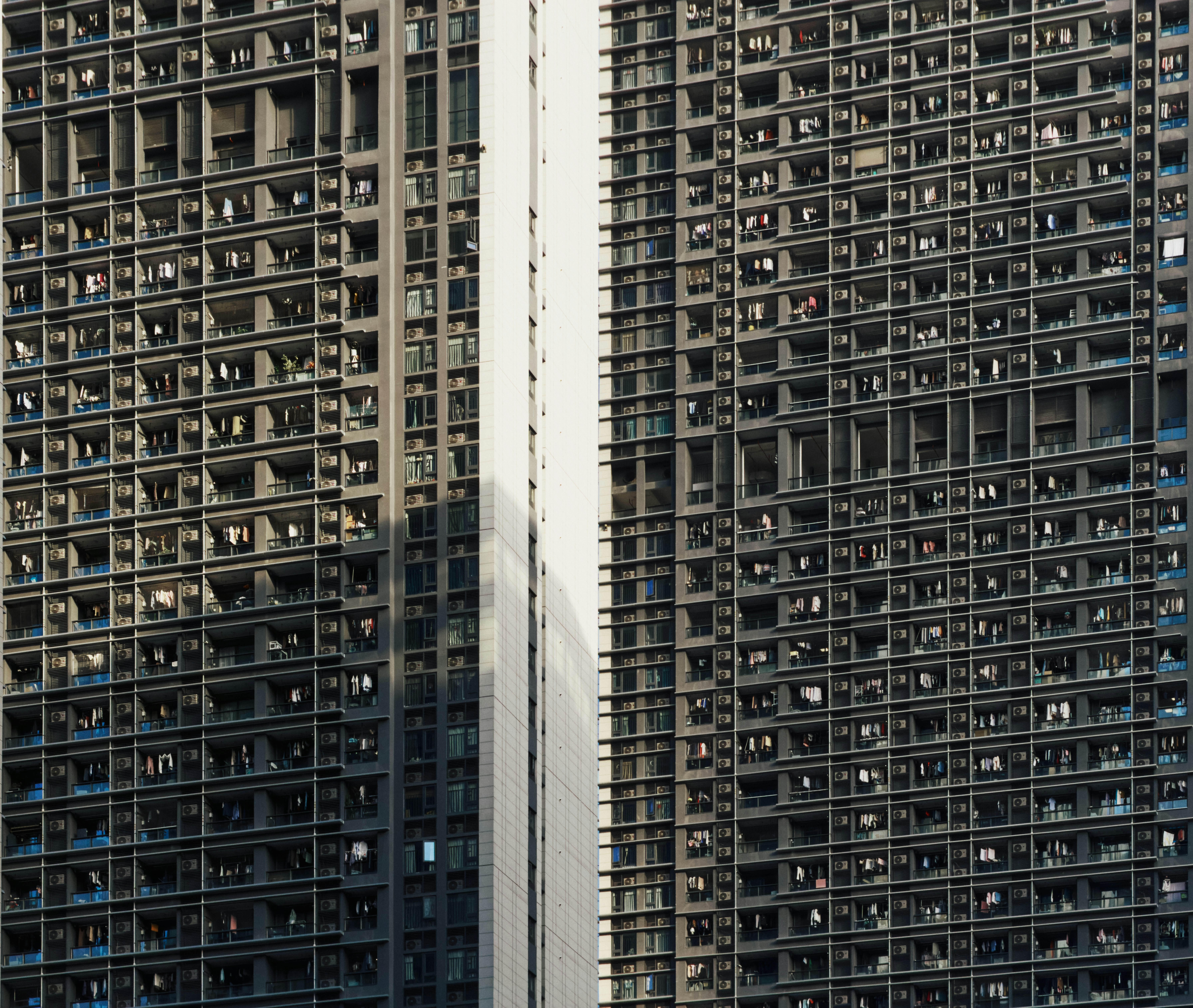 A dense residential high-rise captured during a workday in the city. Repeating balconies, air-conditioning units, and hanging laundry form a rigid grid, revealing traces of everyday life within an overwhelming scale. The symmetry and vertical density reflect the reality of urban living, raising quiet questions about space, privacy, and how it feels to live high above the ground.