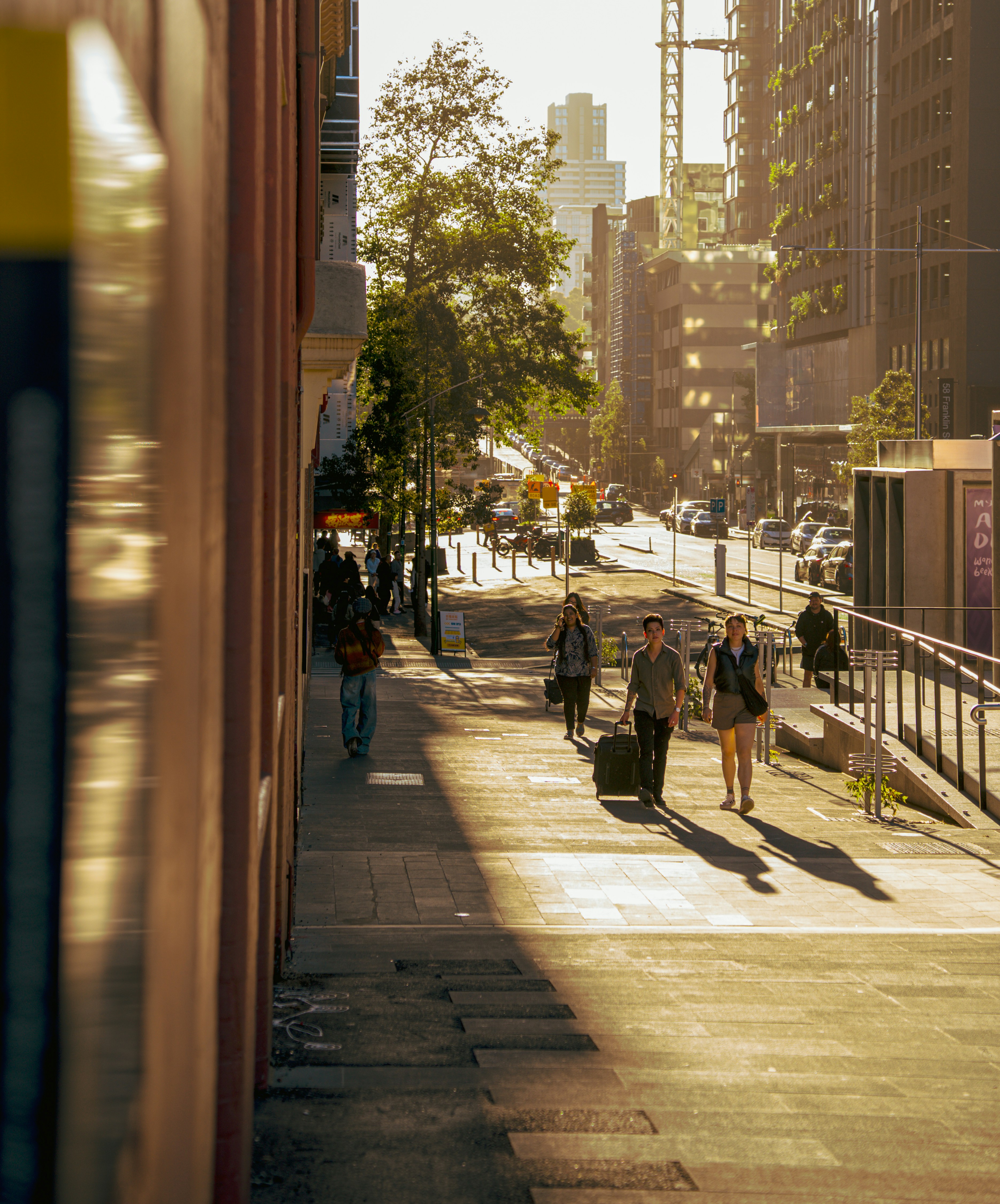 Busy street scene in Melbourne CBD bathed in golden hour light. Pedestrians crossing the road with dramatic long shadows and sun rays cutting through the city air