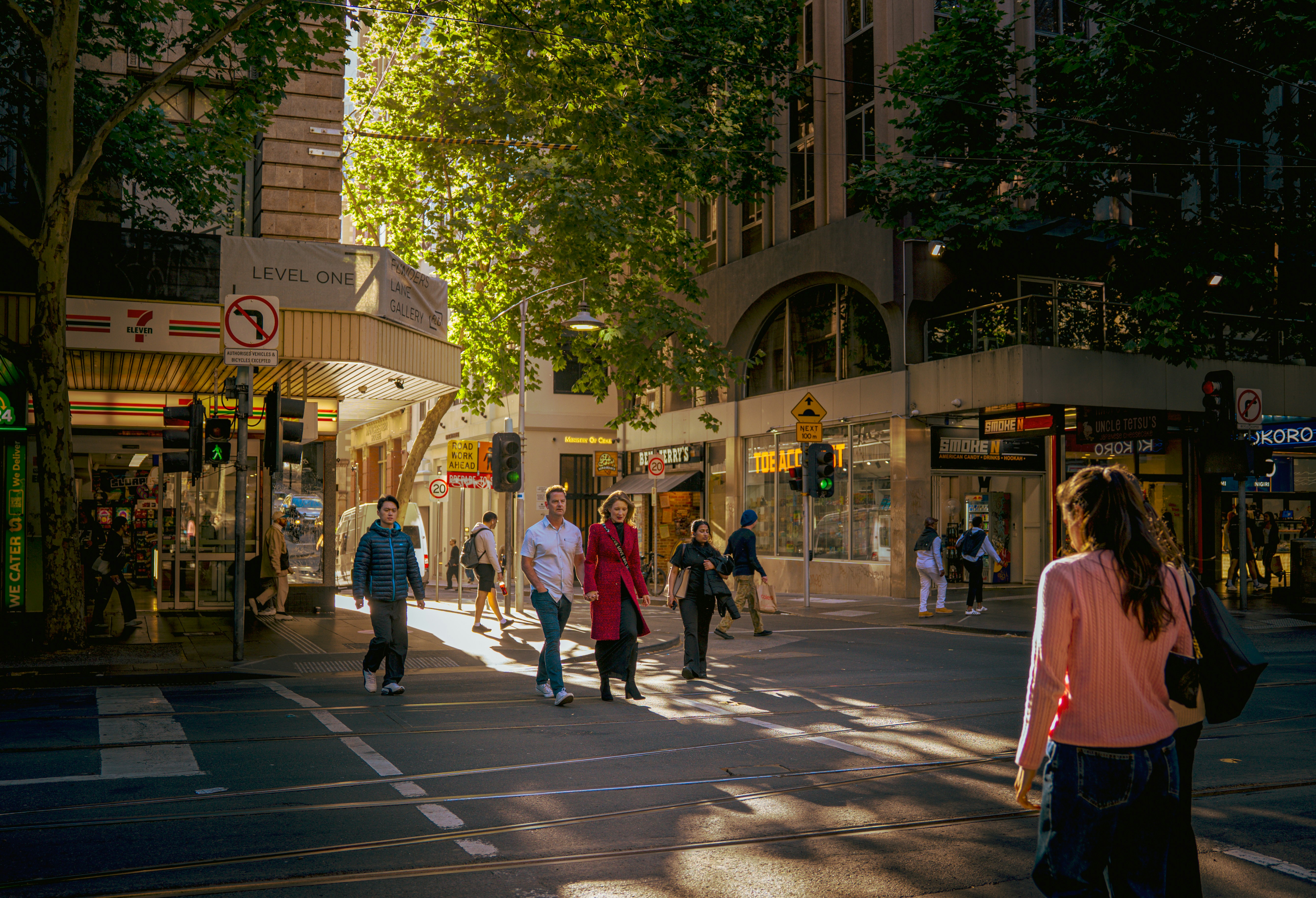 Busy street scene in Melbourne CBD bathed in golden hour light. Pedestrians crossing the road with dramatic long shadows and sun rays cutting through the city air