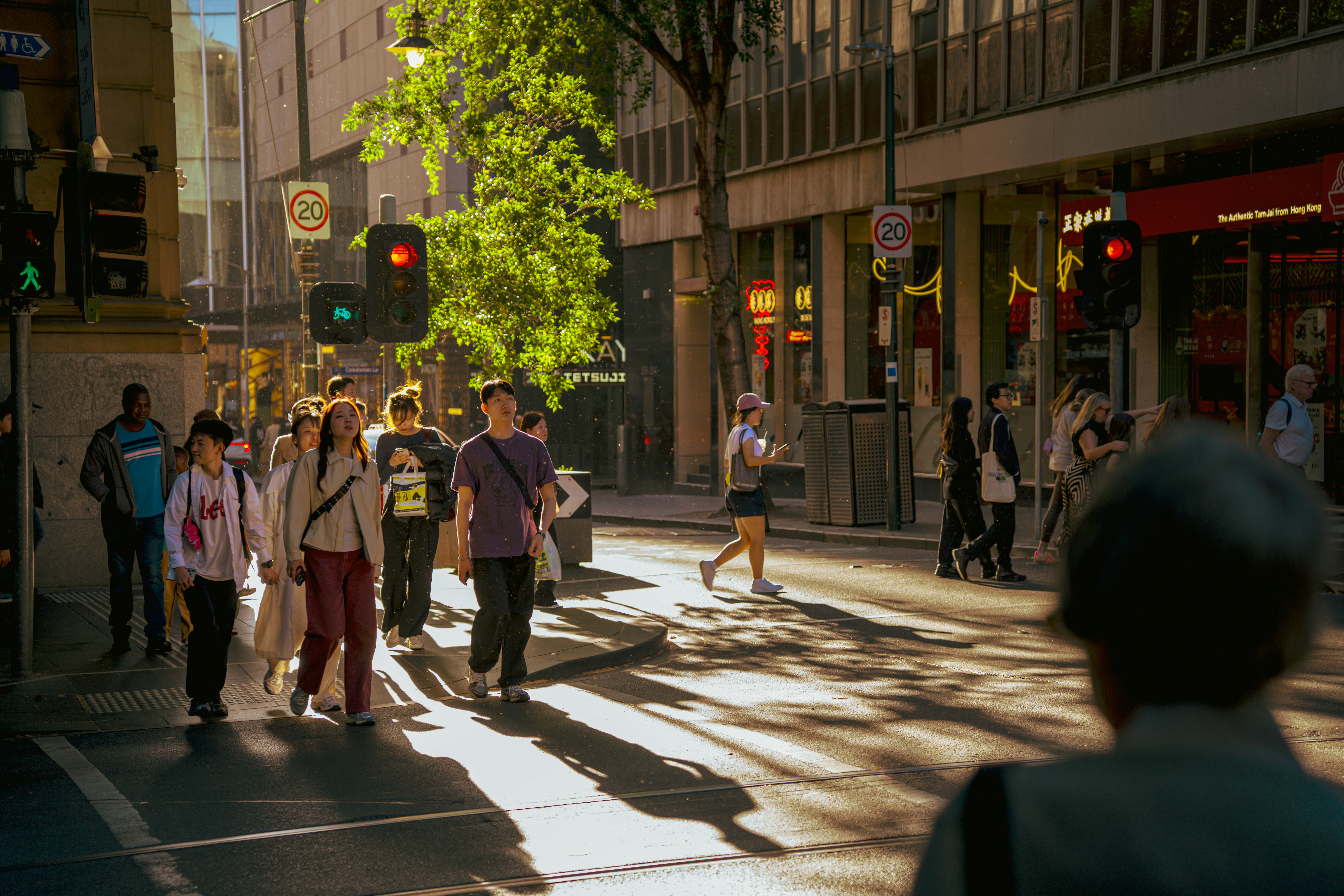 Busy street scene in Melbourne CBD bathed in golden hour light. Pedestrians crossing the road with dramatic long shadows and sun rays cutting through the city air