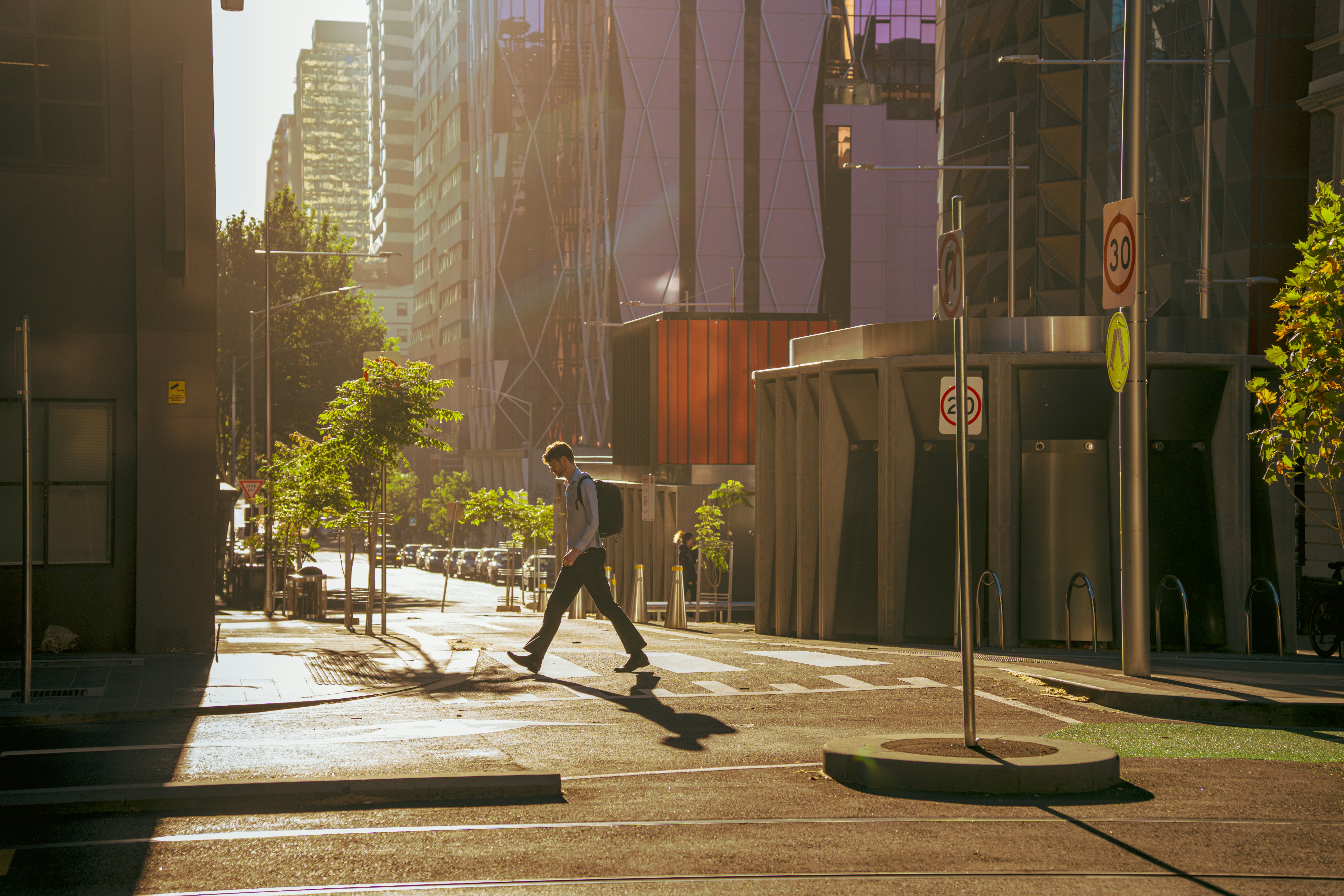 Busy street scene in Melbourne CBD bathed in golden hour light. Pedestrians crossing the road with dramatic long shadows and sun rays cutting through the city air