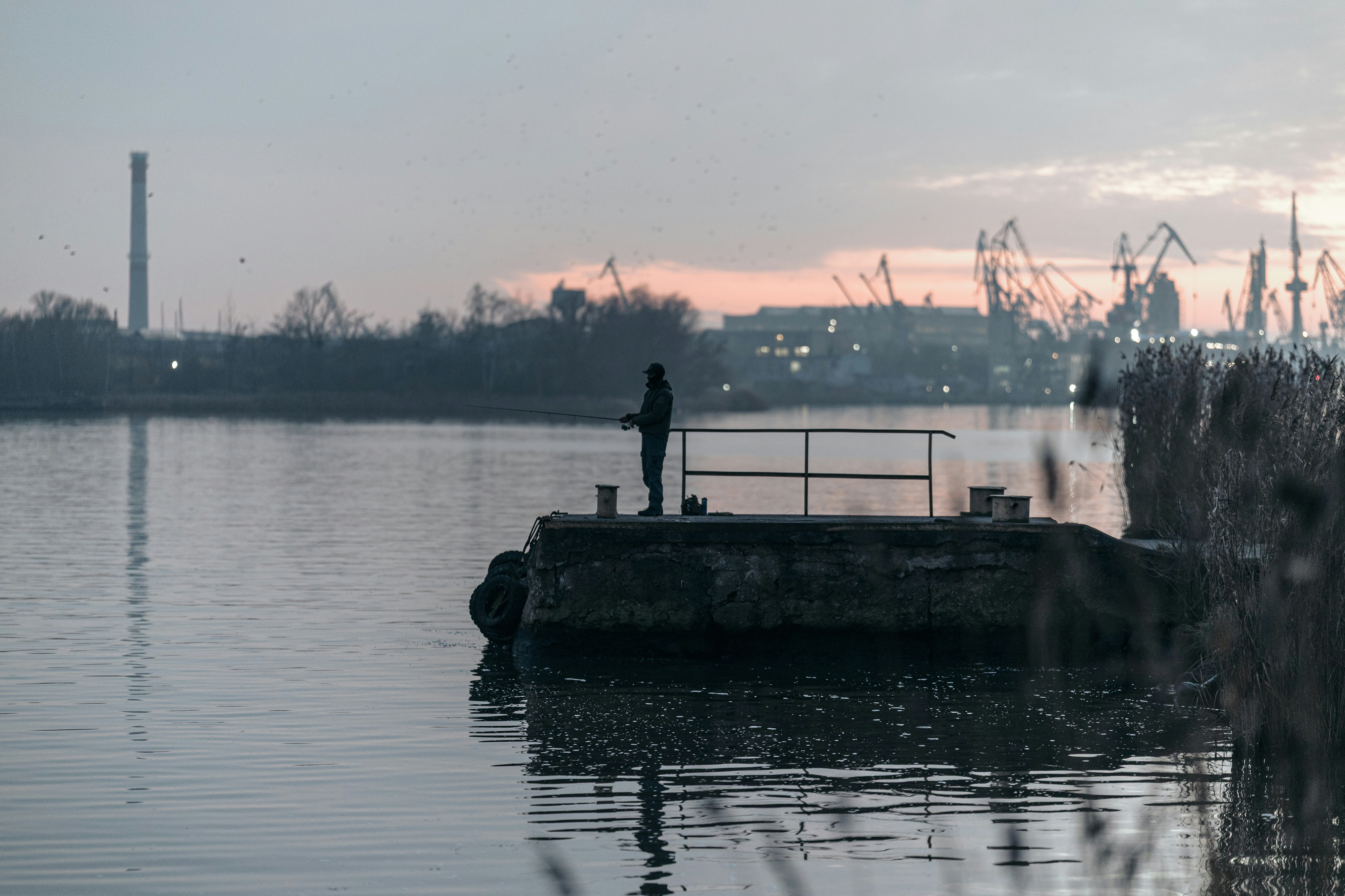 Silhouetted Fisherman at Dusk Along a Quiet Industrial Waterfront