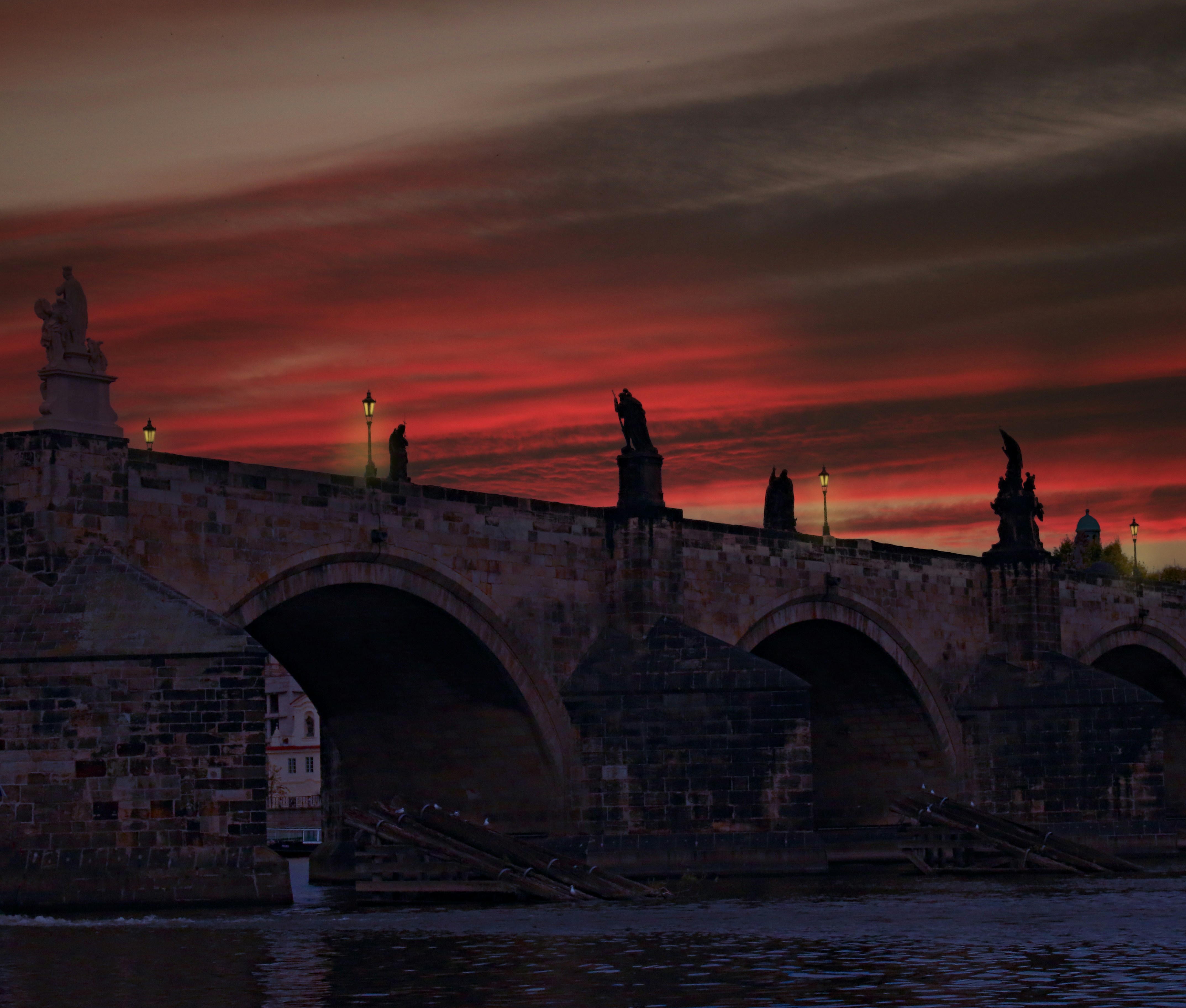 The photograph depicts Charles Bridge in Prague silhouetted against a dramatic sunset, with the sky ablaze in vivid shades of red and black. The statues adorning the bridge stand in stark silhouette, creating a striking contrast with the evening light. The scene evokes mystery, history, and an almost theatrical beauty over the Vltava River.