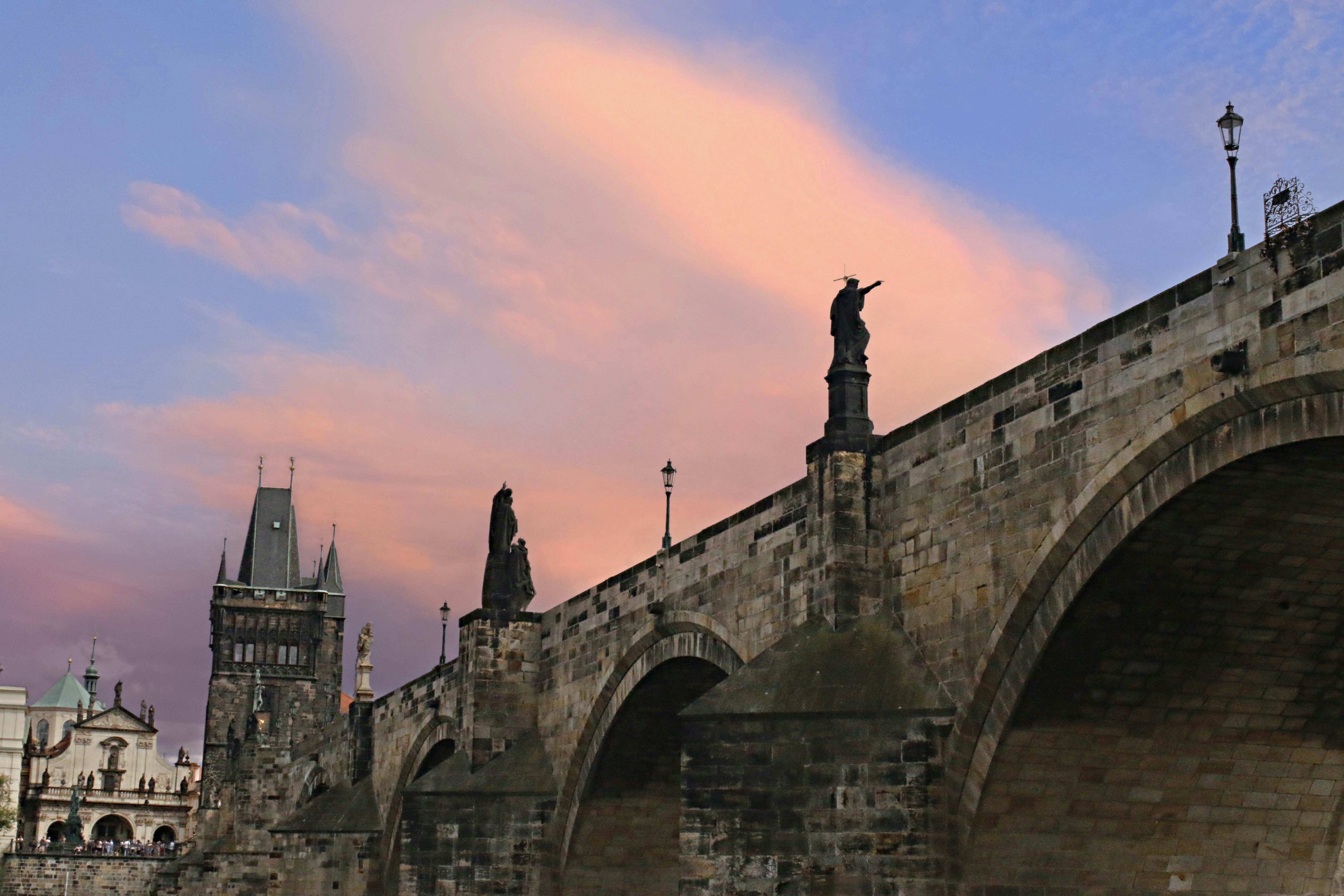 The photograph captures the historic Charles Bridge, one of Prague’s most iconic landmarks, bathed in the warm hues of sunset. The baroque statues rise majestically above the stone arches, while the sky, painted in shades of pink and blue, envelops the scene with a romantic and timeless atmosphere.