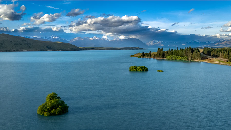 A tranquil lakescape with small tree-covered islets and distant mountains under a bright blue sky; a wide-angle view emphasizing expansive water and shoreline detail. (Drone-style landscape photo).